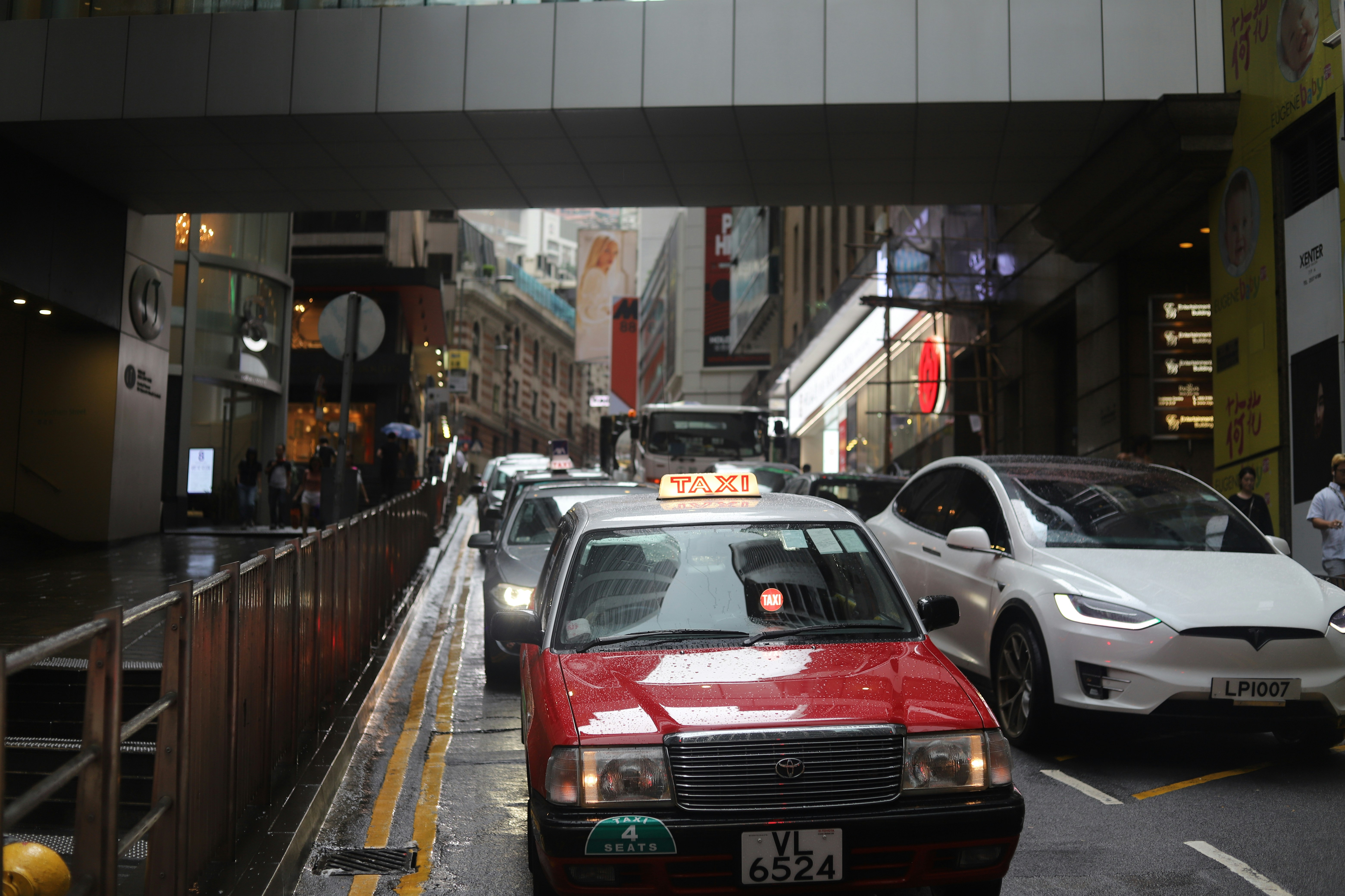A red taxi navigates a busy street in a bustling city, surrounded by modern vehicles and urban architecture. Raindrops glisten on the pavement.