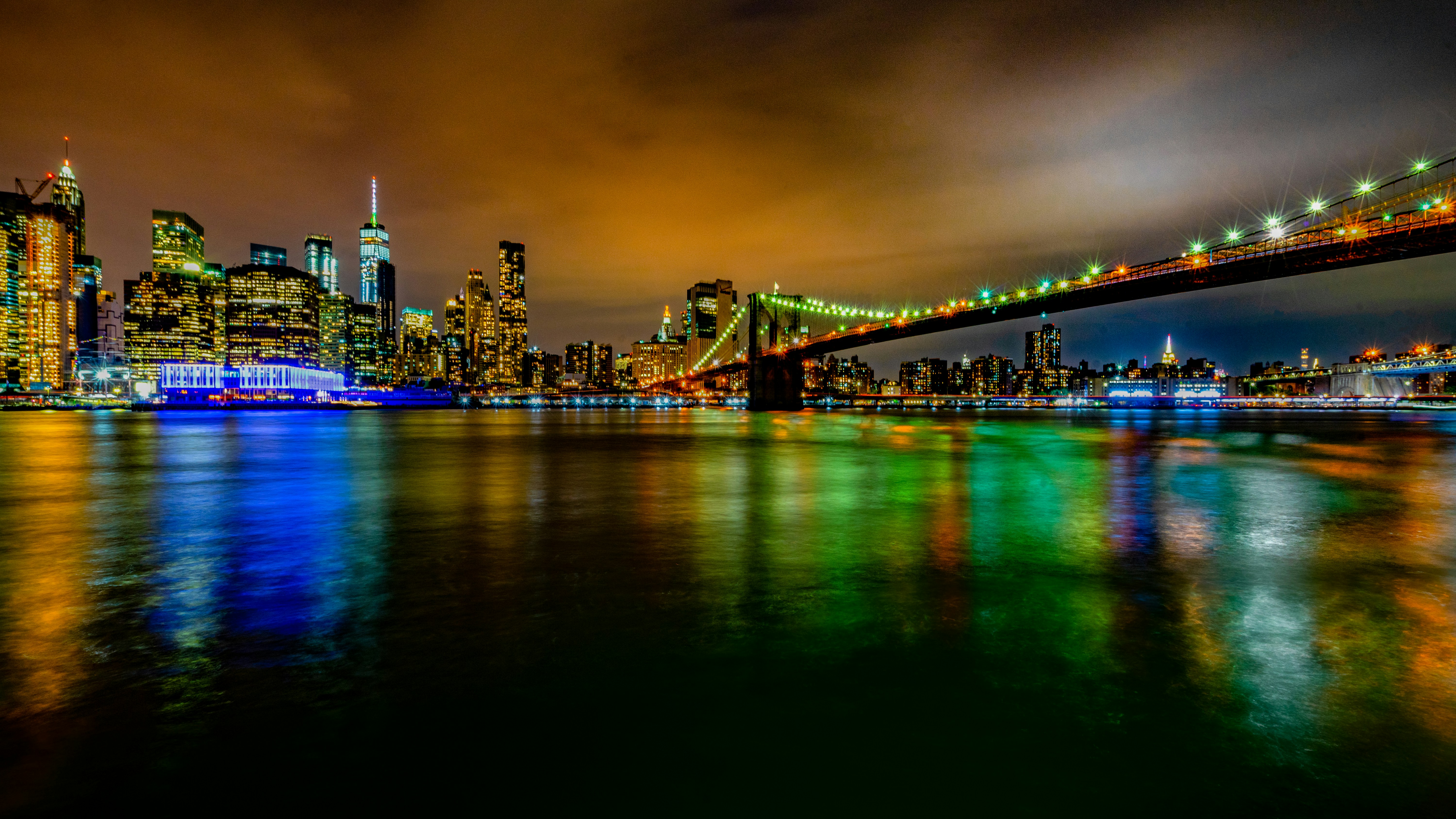 Vibrant city skyline illuminated at night, showcasing the Brooklyn Bridge and colorful reflections on the water's surface.