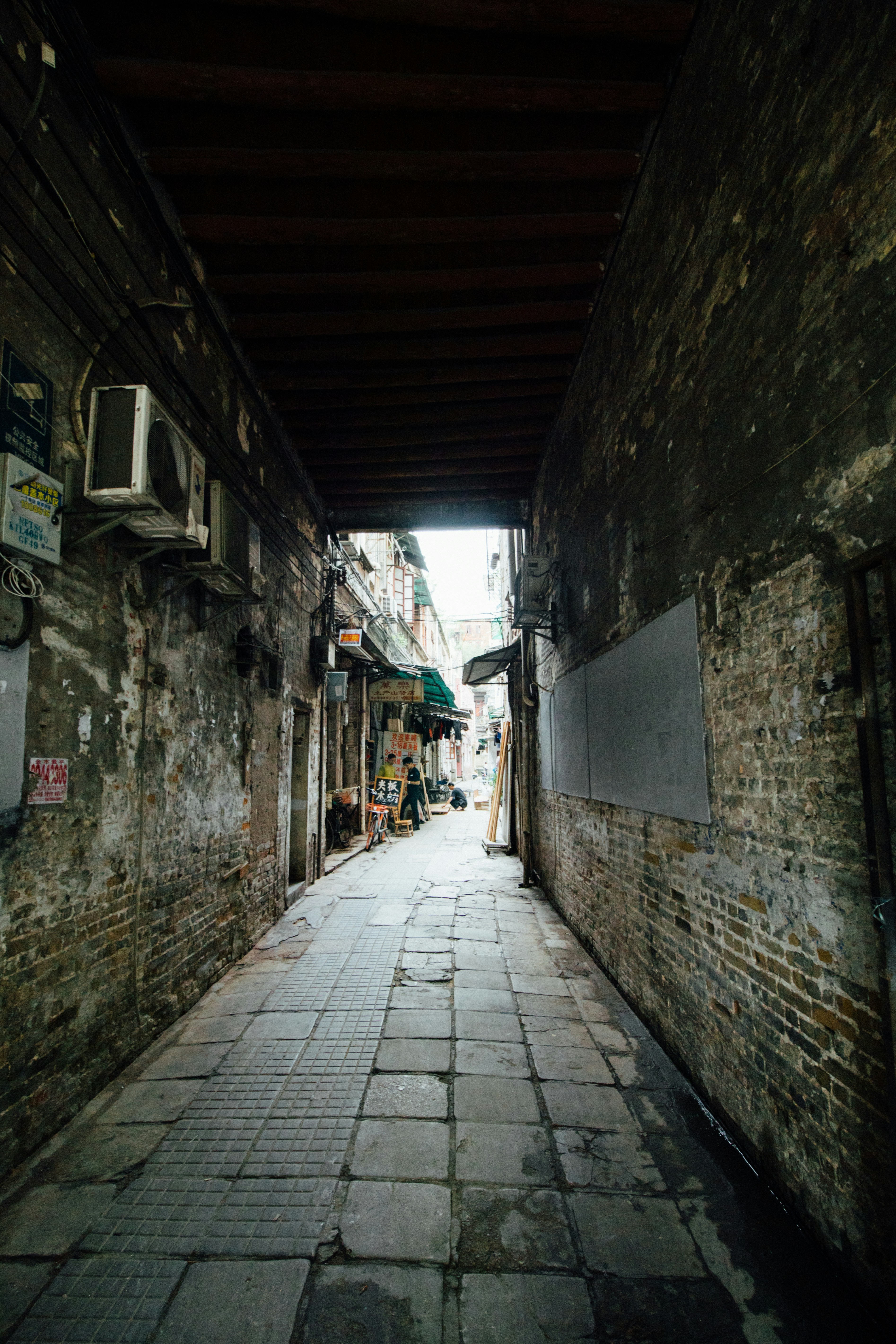 Narrow alleyway flanked by weathered brick walls and old signage, leading to a distant opening. The scene captures a blend of urban decay and quiet charm.