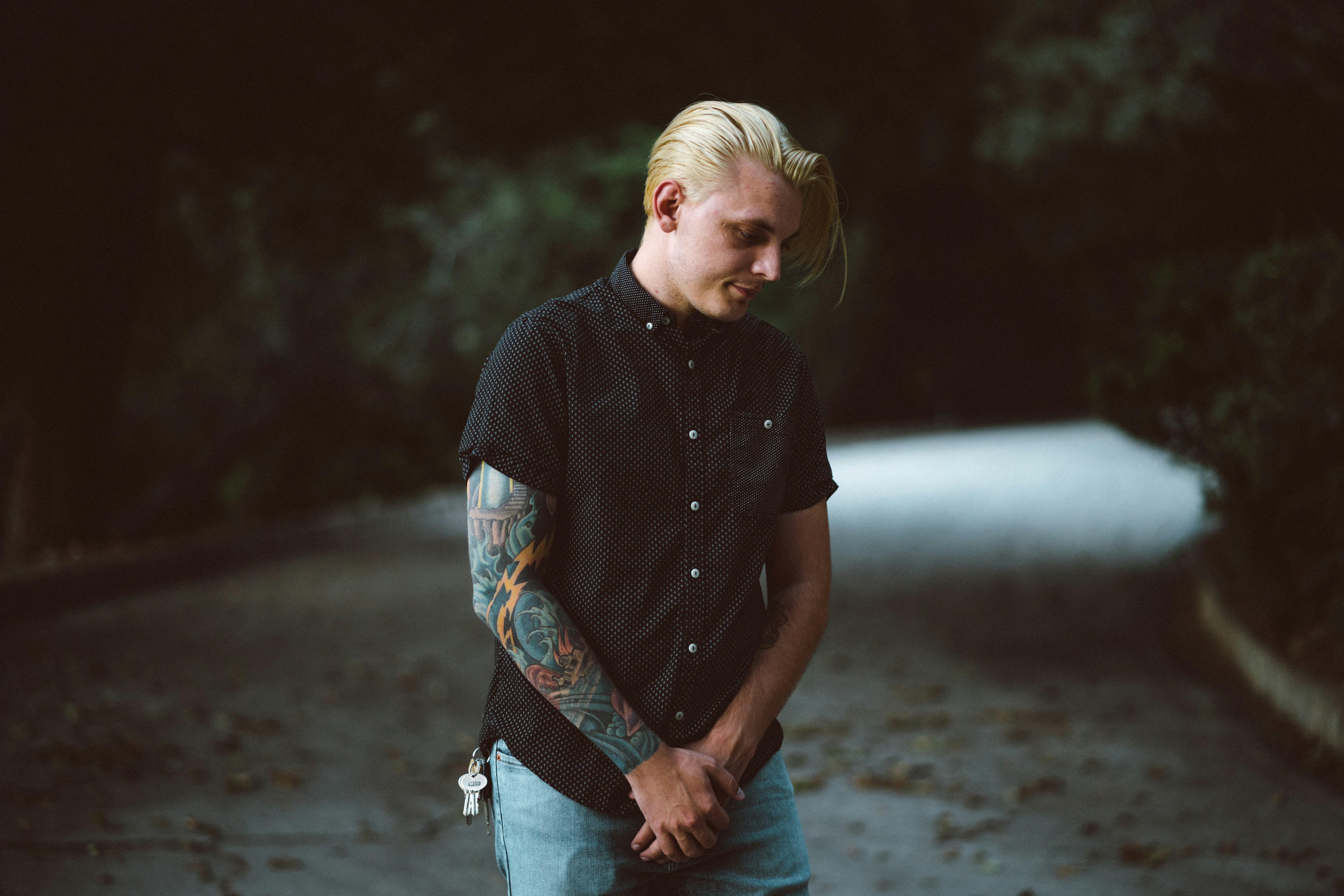 Young man with blonde hair and tattoos stands contemplatively on a dimly lit path, embodying a moment of introspection.
