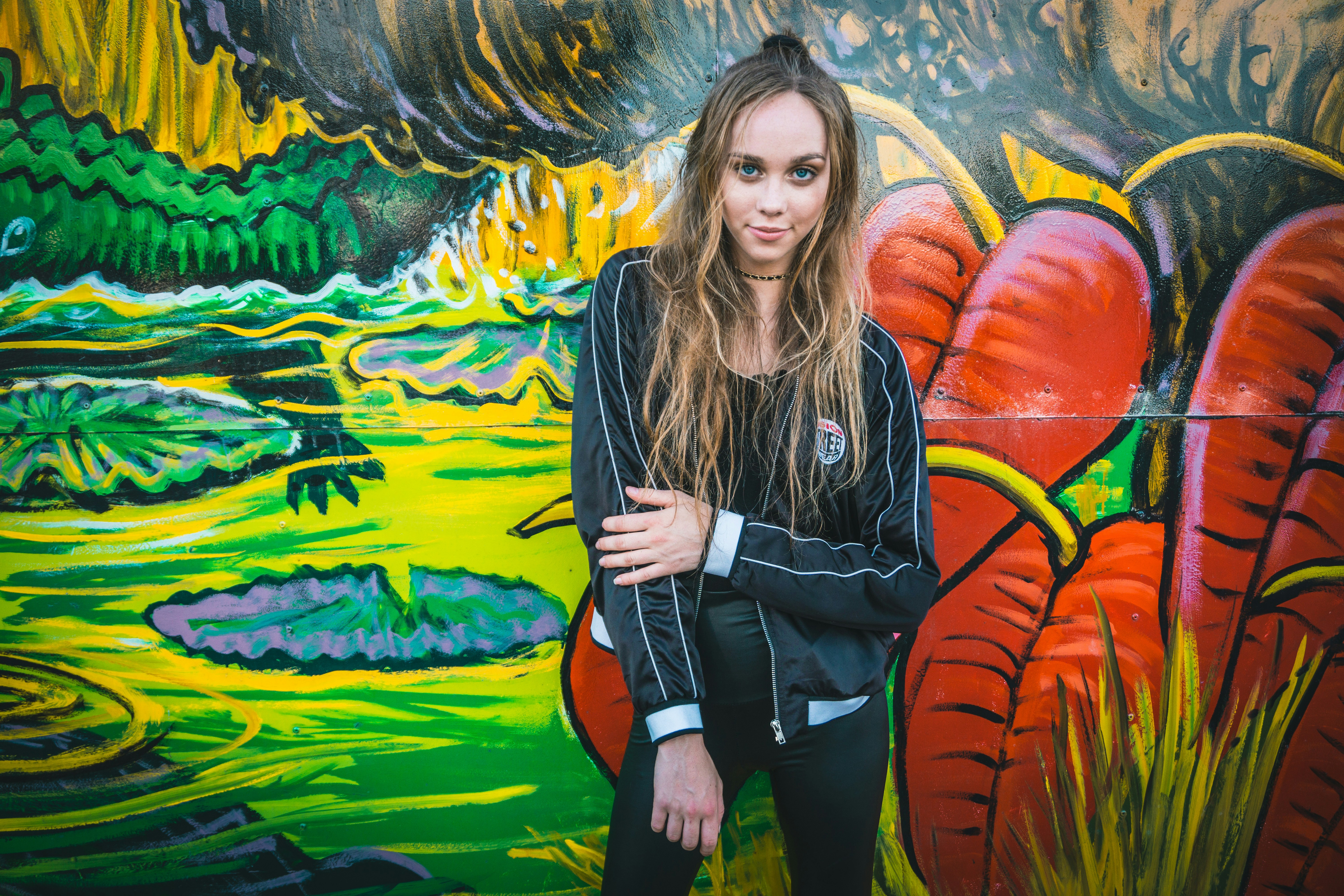 A young woman poses confidently in front of a vibrant mural featuring lush greenery and bold colors, embodying the spirit of urban art.