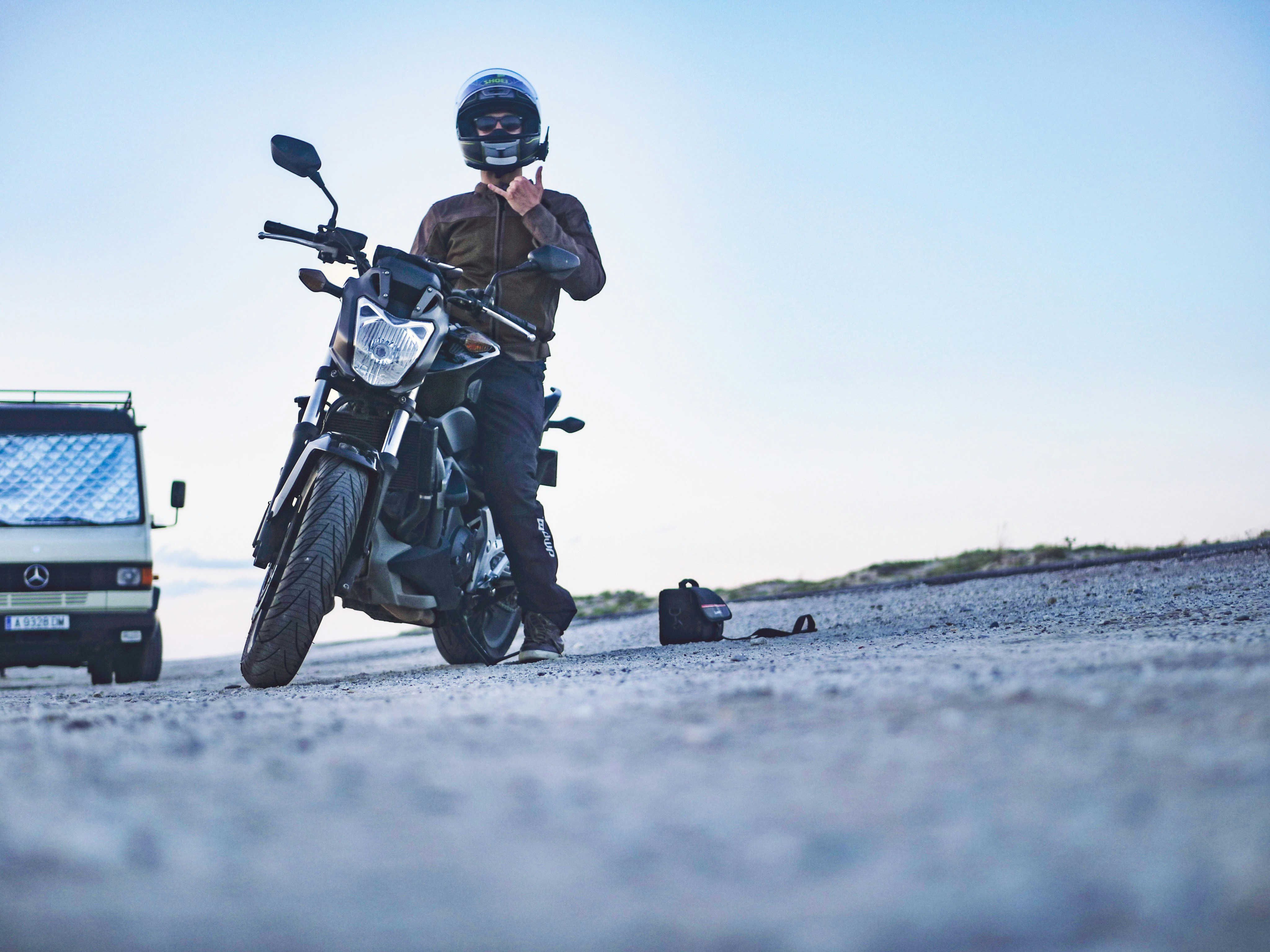 Motorcyclist adjusting helmet while standing beside a bike on an open road, with a van in the background. 