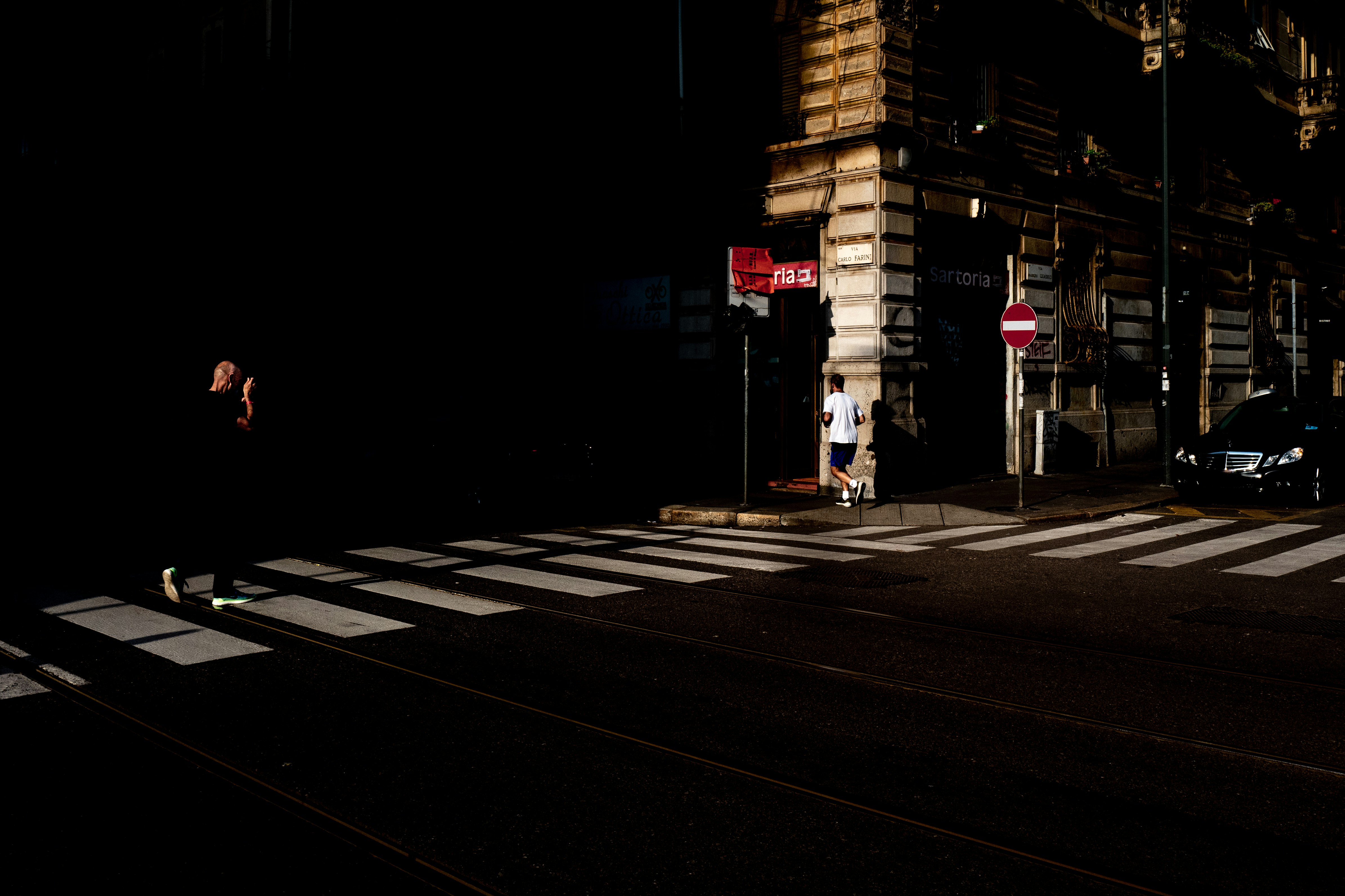 A pedestrian crossing illuminated by strong sunlight, contrasting sharply with the surrounding shadows. The scene captures the urban rhythm of city life.