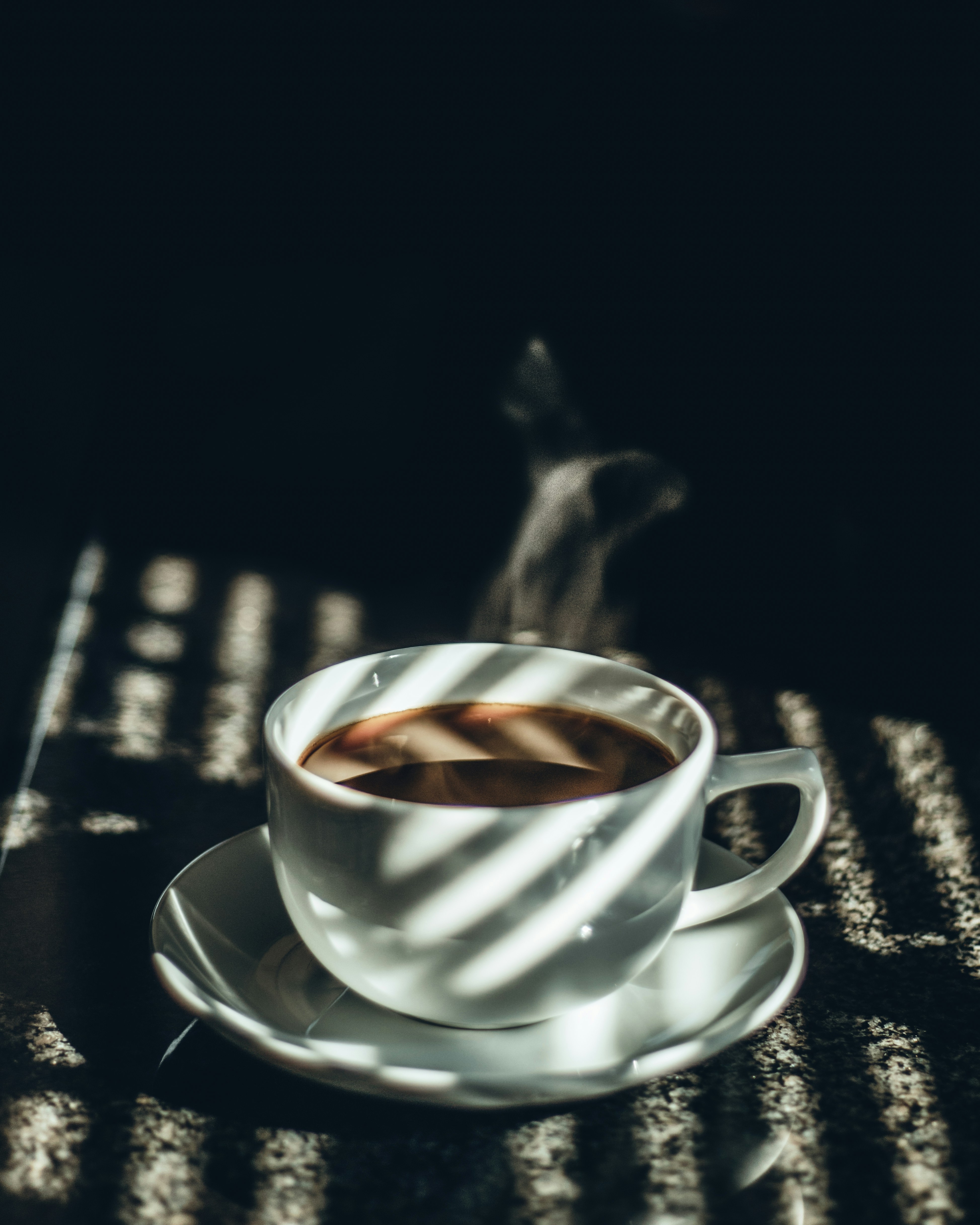 White coffee cup resting on a plate, surrounded by dramatic shadows and light patterns. Rich coffee fills the cup, creating a warm focal point.