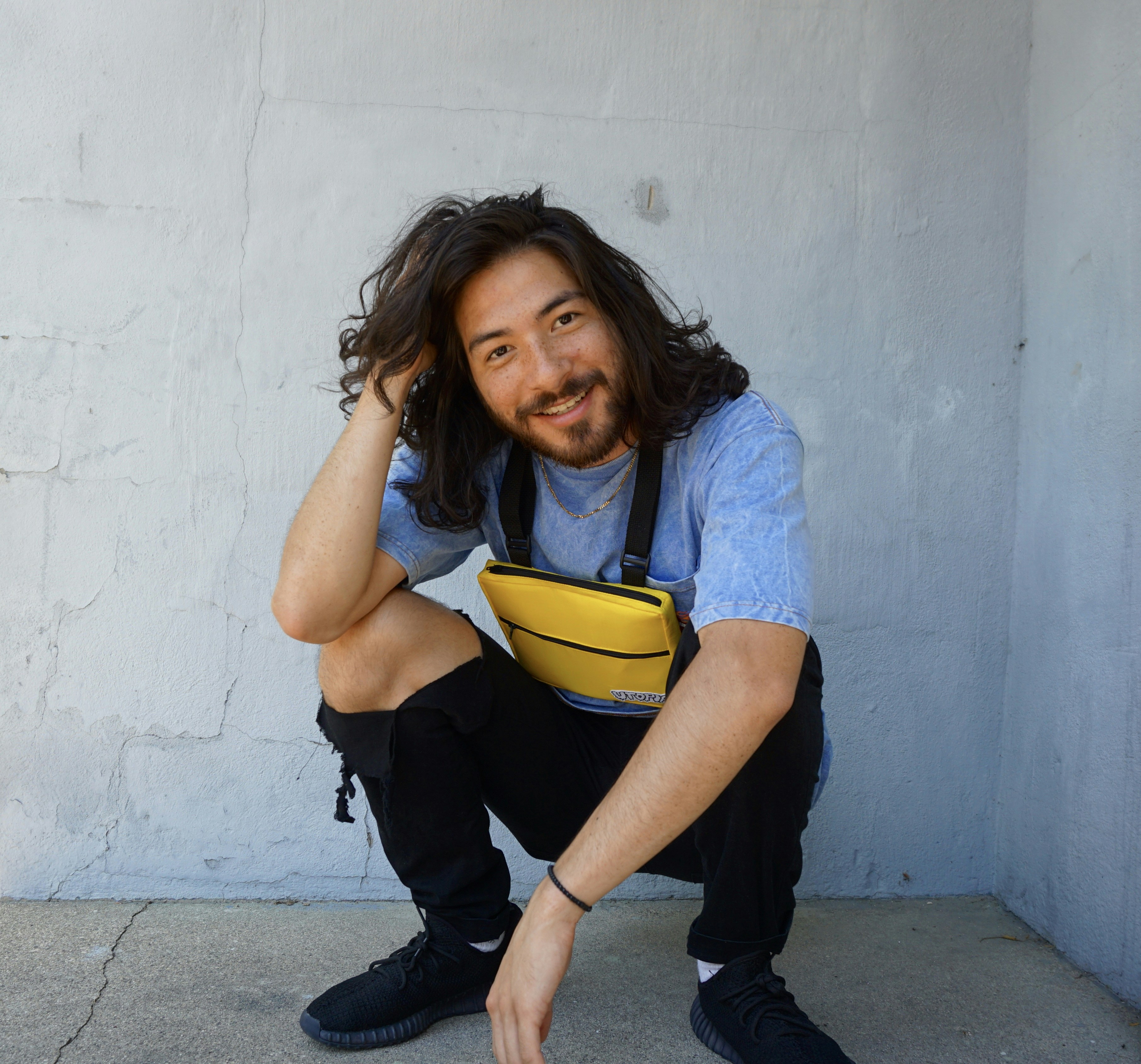 Man in casual attire crouching against a textured wall, wearing a yellow chest rig.