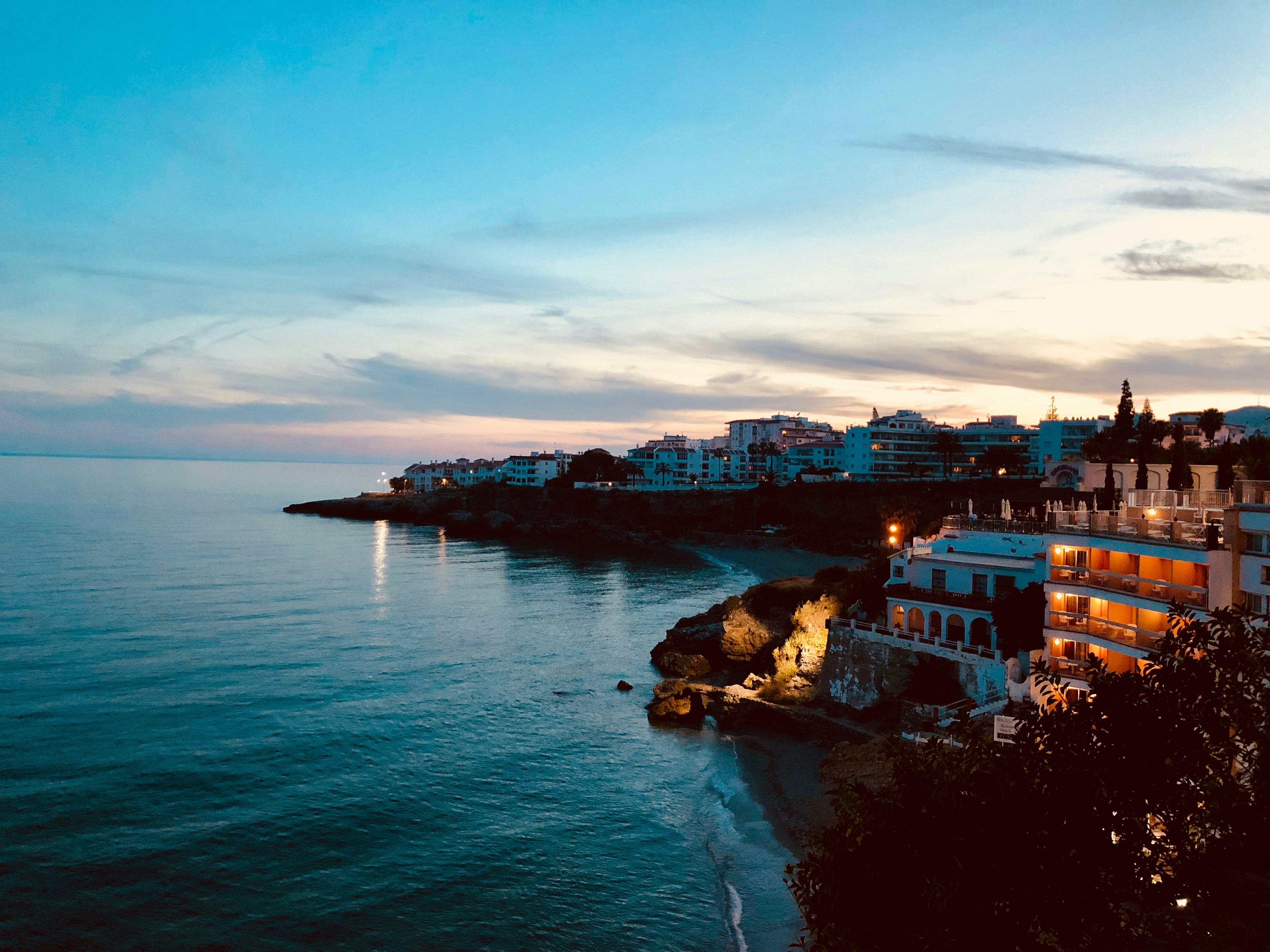 Coastal town at dusk with buildings illuminated along the shoreline and a tranquil sea reflecting the evening sky.