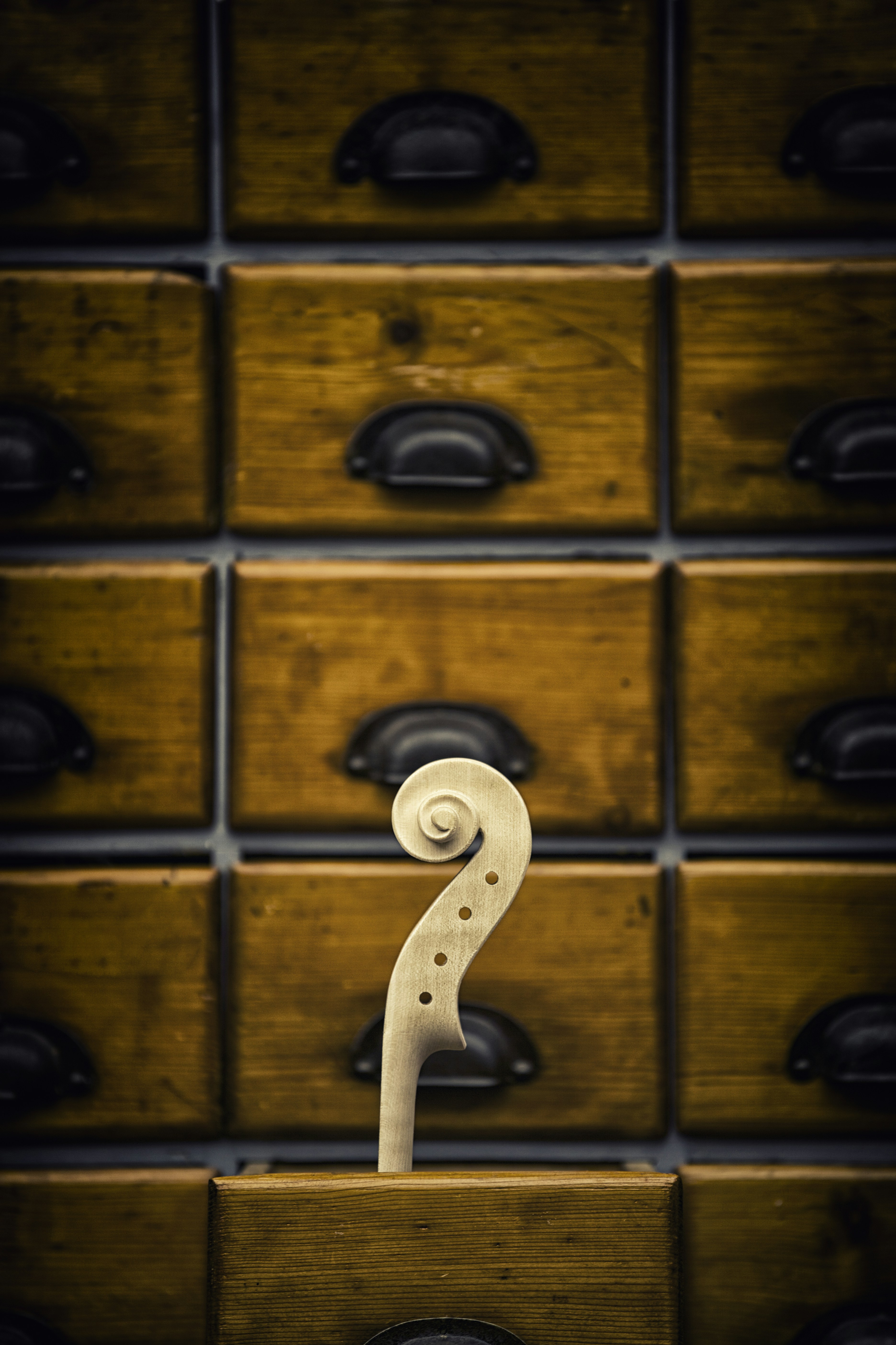 Intricate scroll of a violin resting against a backdrop of wooden drawers, highlighting the artistry of instrument making.