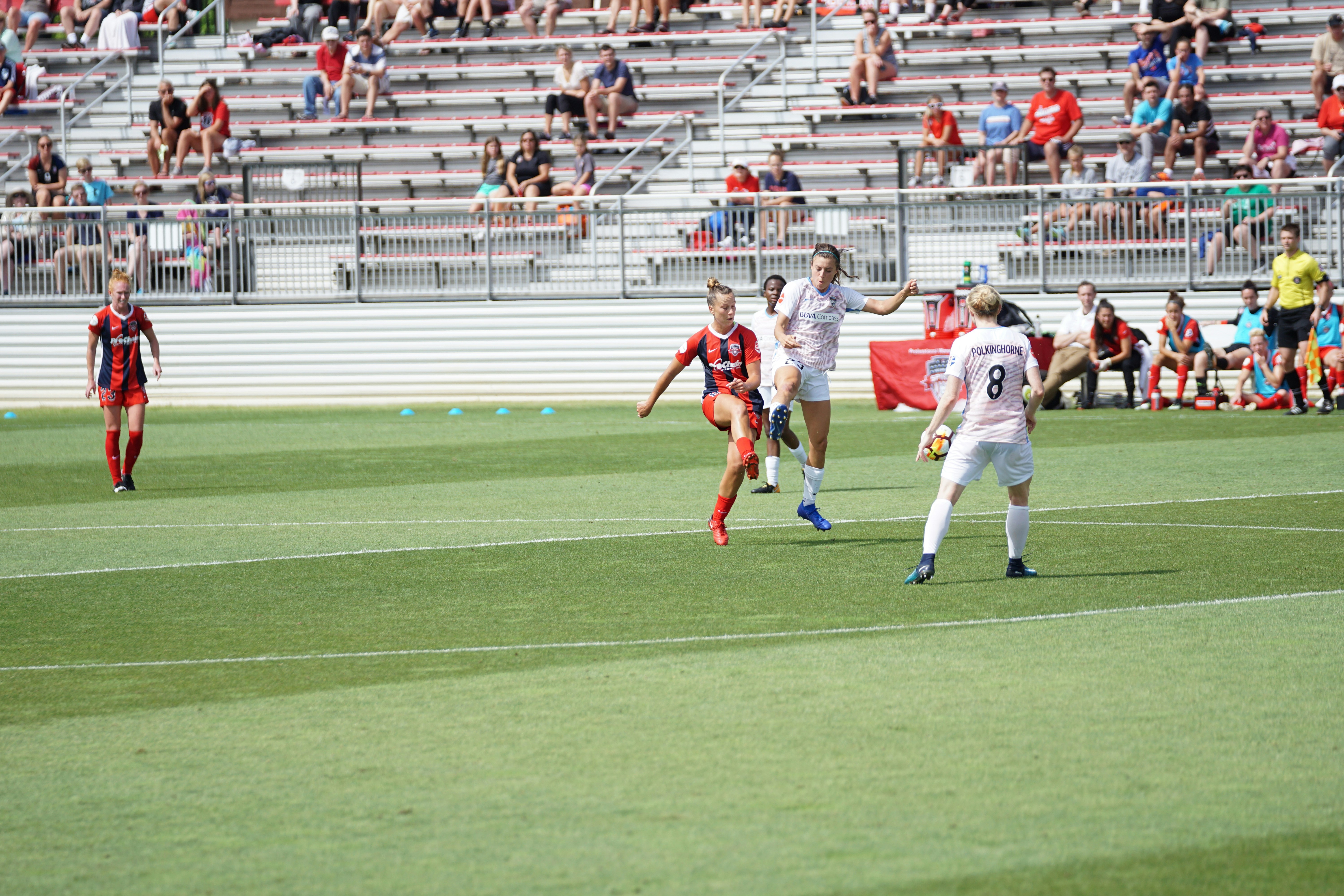 Soccer players in red and white jerseys compete intensely on a sunny field with a packed stadium in the background.