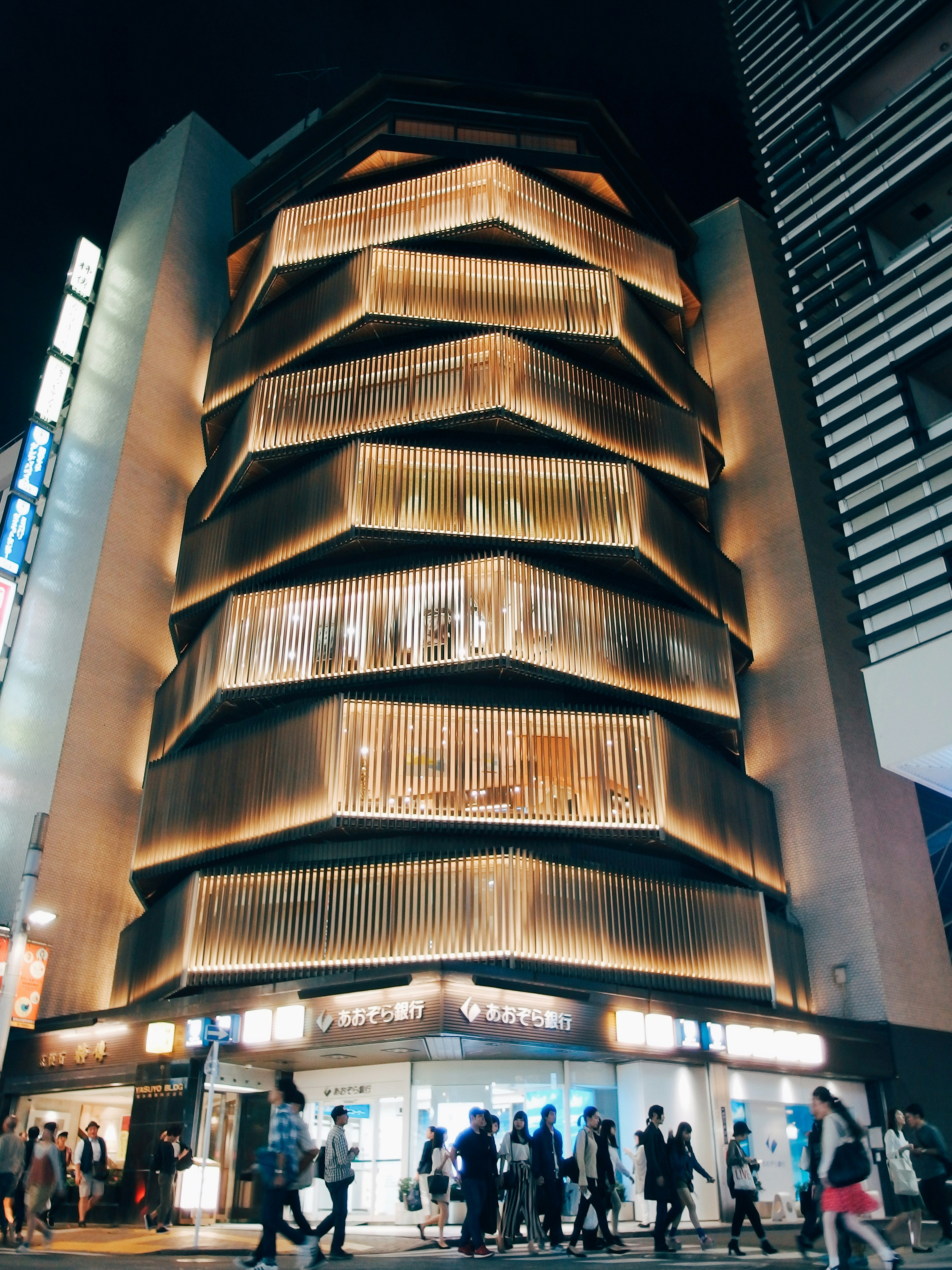 Modern architectural building with layered wooden façade illuminated at night, surrounded by pedestrians. The design showcases a blend of traditional and contemporary styles.