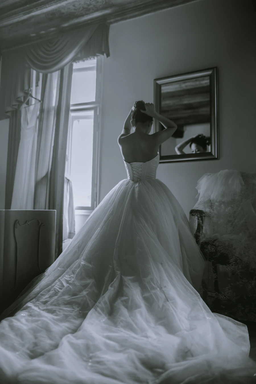 A bride in a voluminous wedding dress stands with her back to the camera, adjusting her hair beside a bright window and wall mirror. The gown’s long train spreads across the room in soft folds.