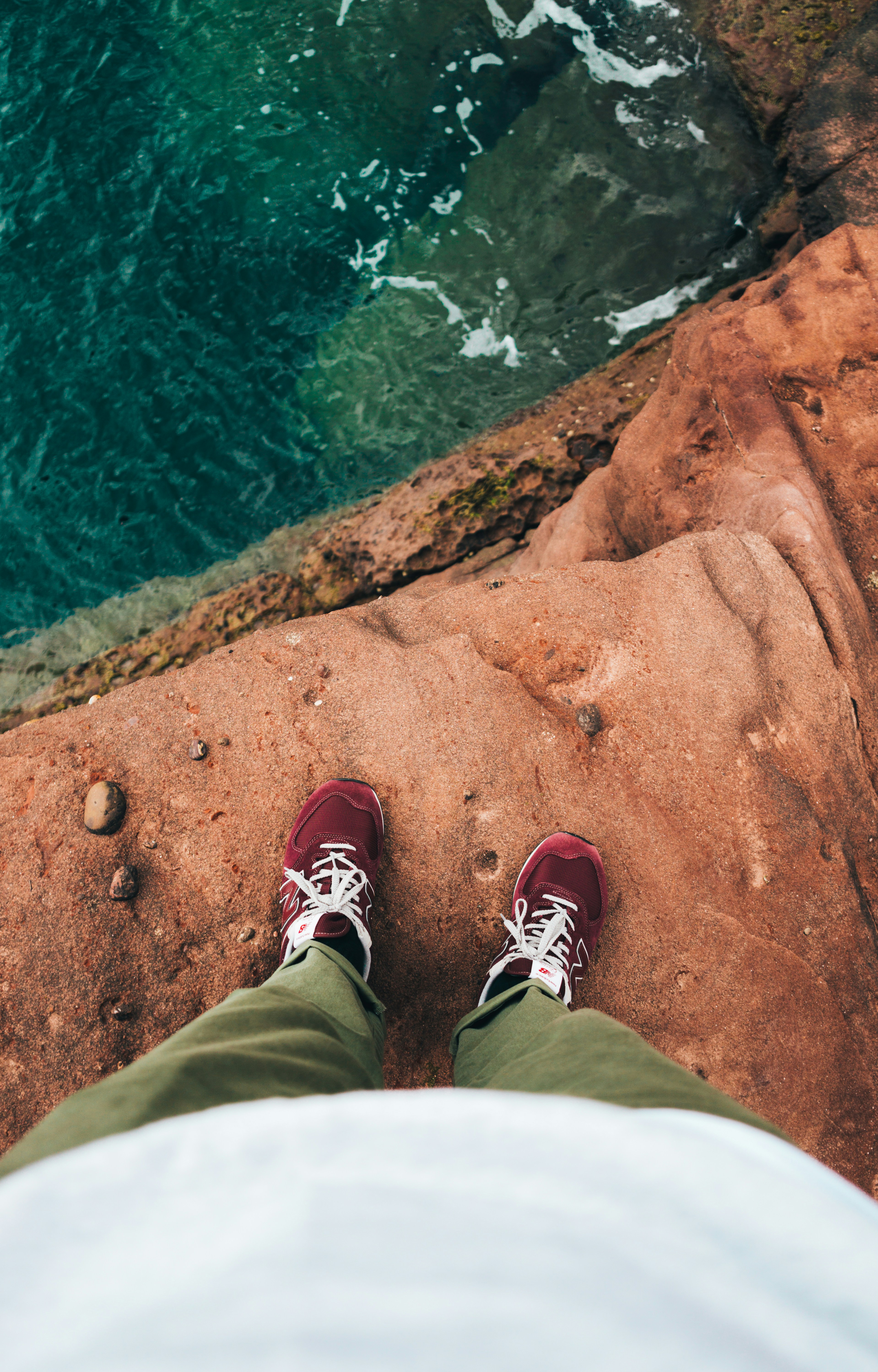 Person standing on a rocky ledge overlooking turquoise waters, with shoes prominently displayed. The scene captures the thrill of nature's beauty and the allure of adventure.