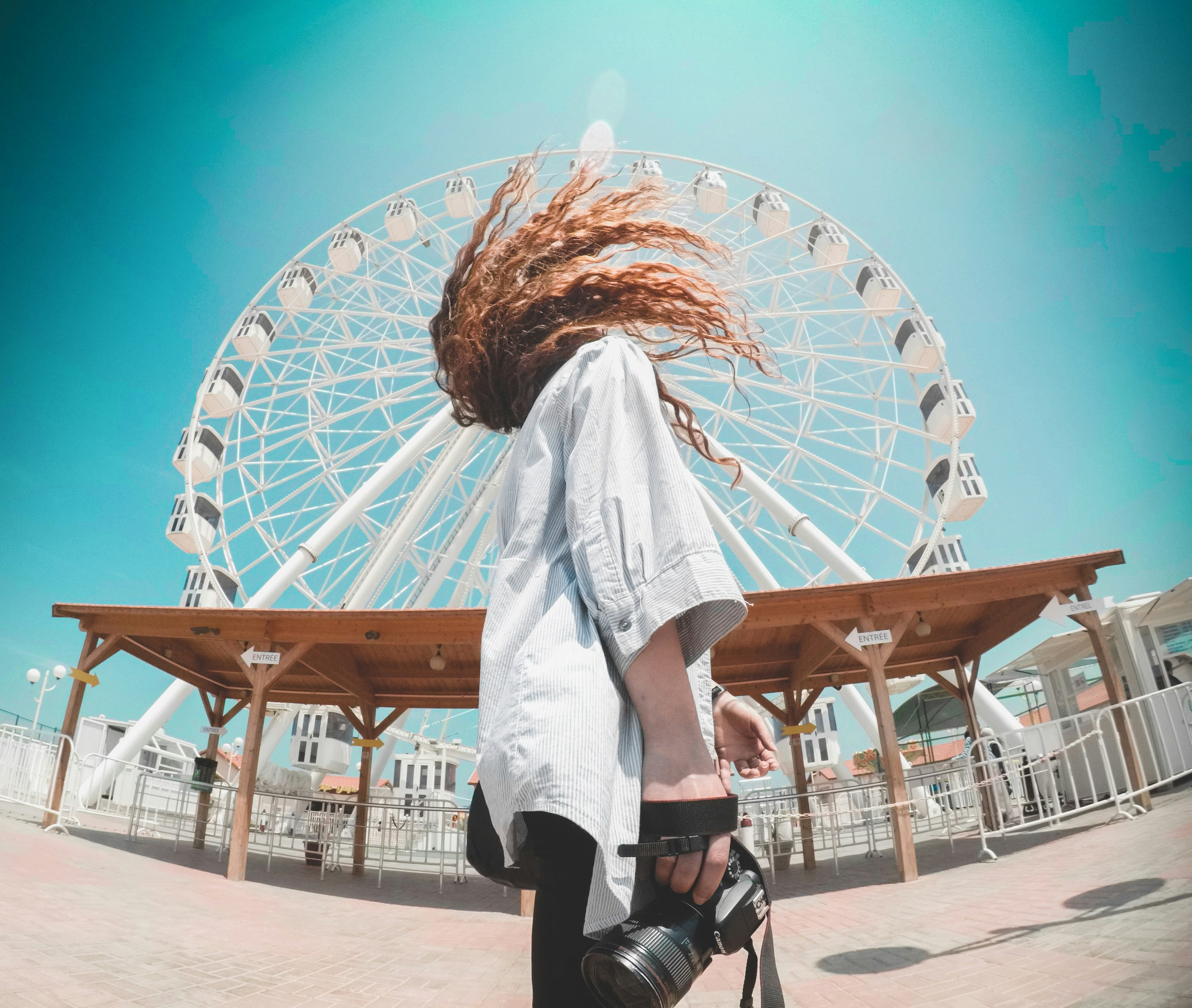 A person with flowing hair stands confidently, holding a camera, framed by a towering Ferris wheel in a vibrant blue sky.
