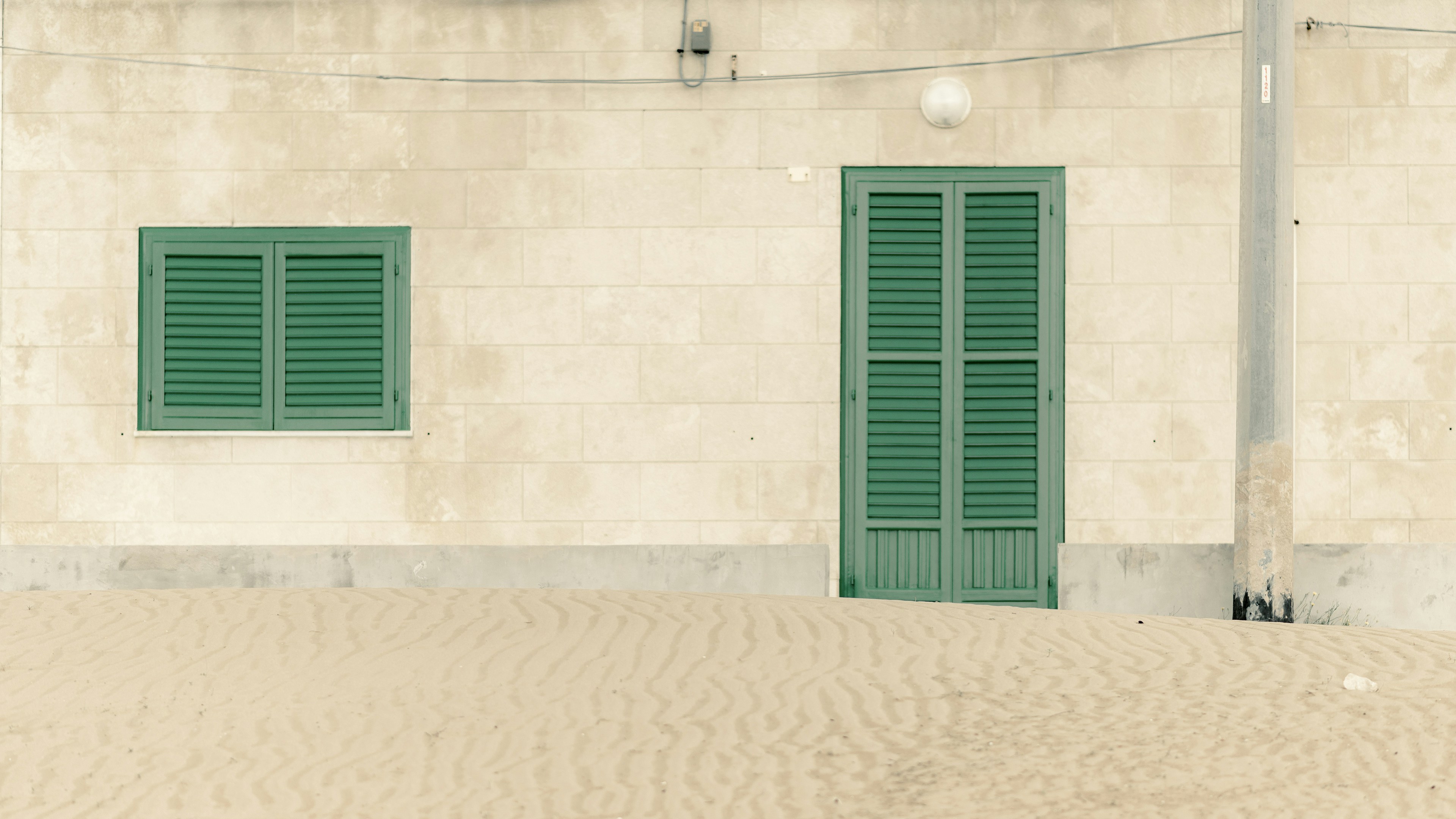 Green-shuttered windows and a door on a weathered wall, partially obscured by shifting sands, evoke a sense of abandonment. The scene captures the interplay between architecture and nature's encroachment.