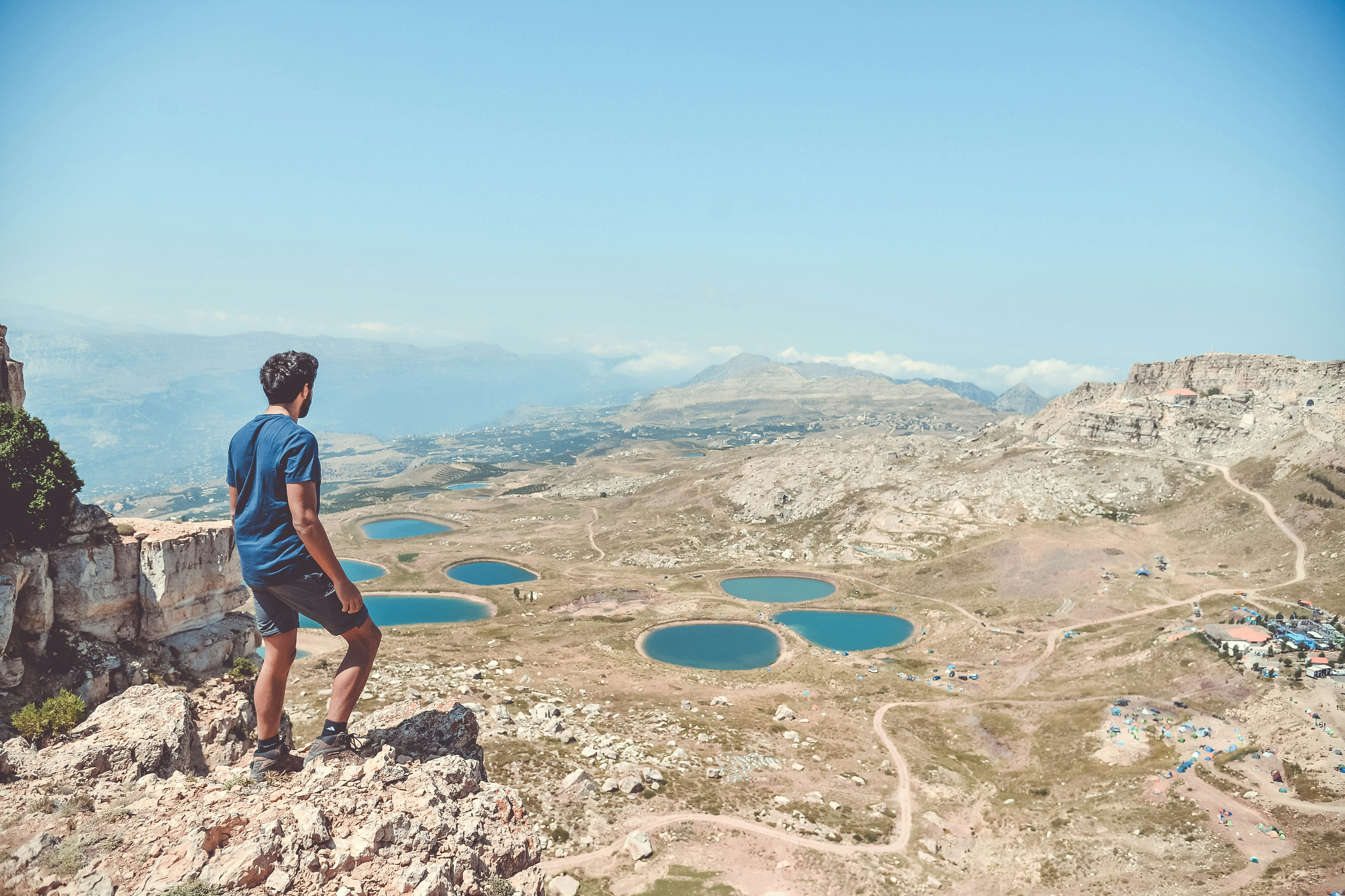 Person standing on rocky cliff gazing at multiple blue lakes in a vast, rugged landscape under a clear sky.