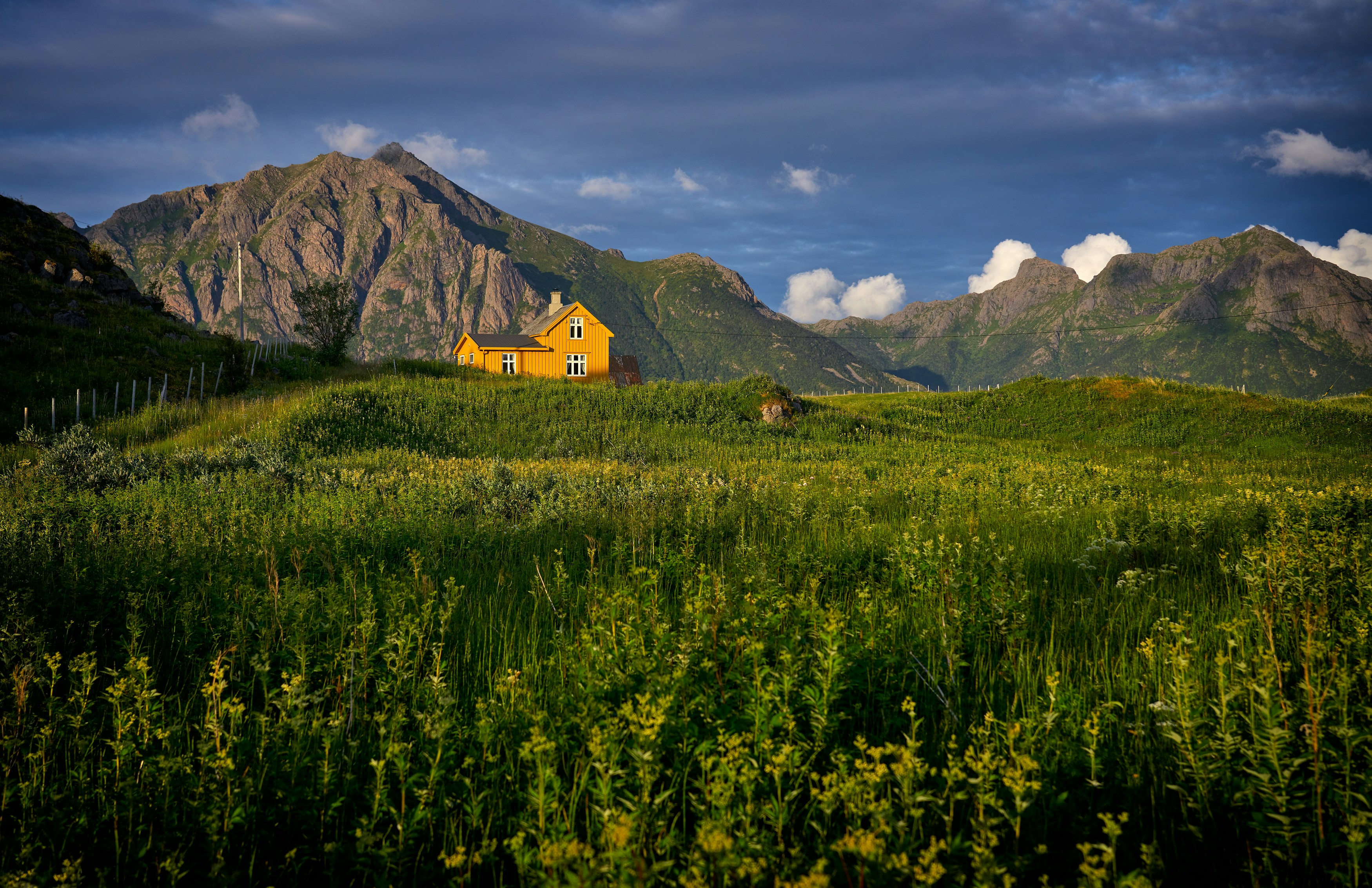 Bright yellow house on a lush green hillside under dramatic mountain backdrop.
