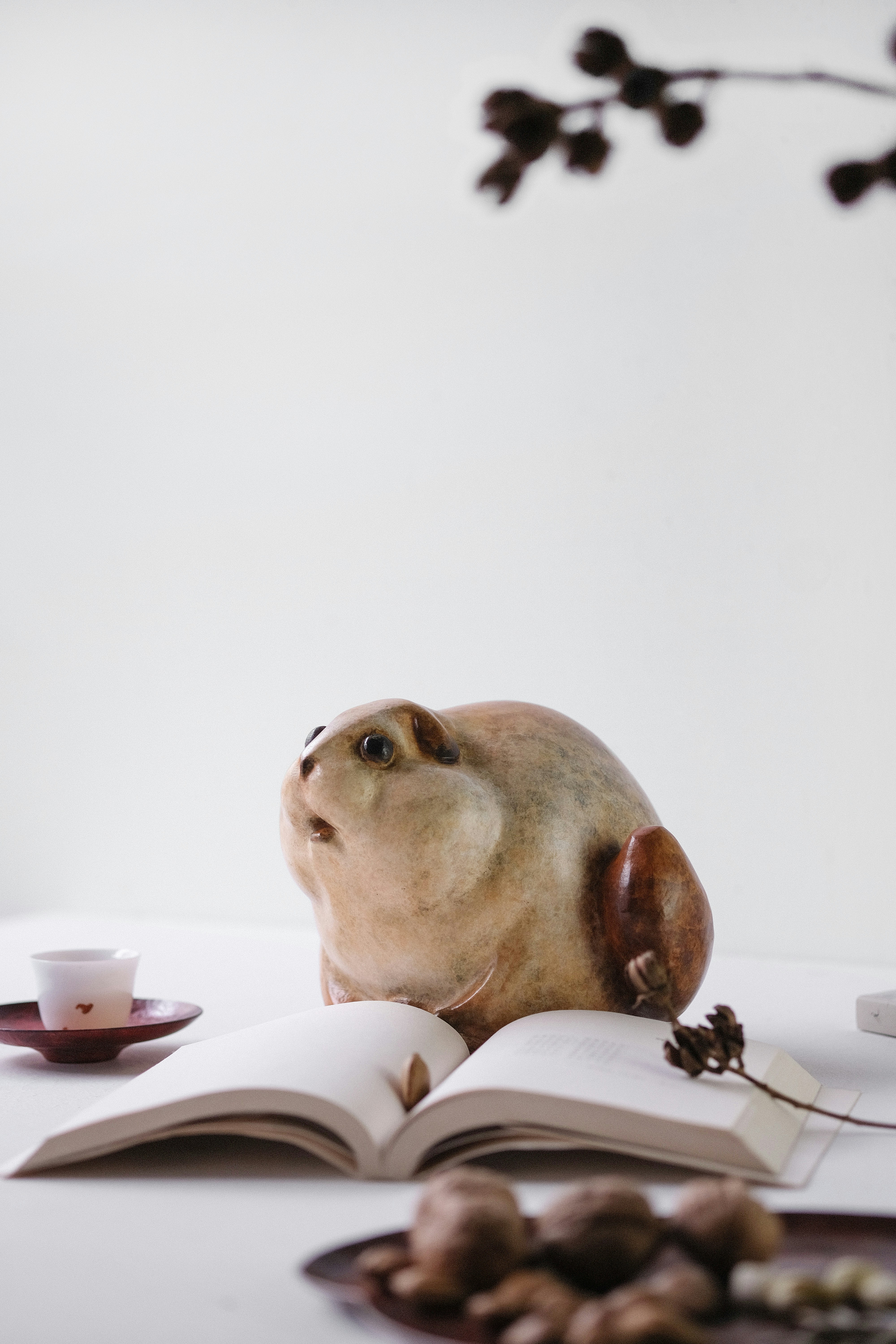 A whimsical ceramic creature resembling a round rabbit sits beside an open book and delicate tea set, surrounded by natural elements. The composition evokes a sense of quiet contemplation.
