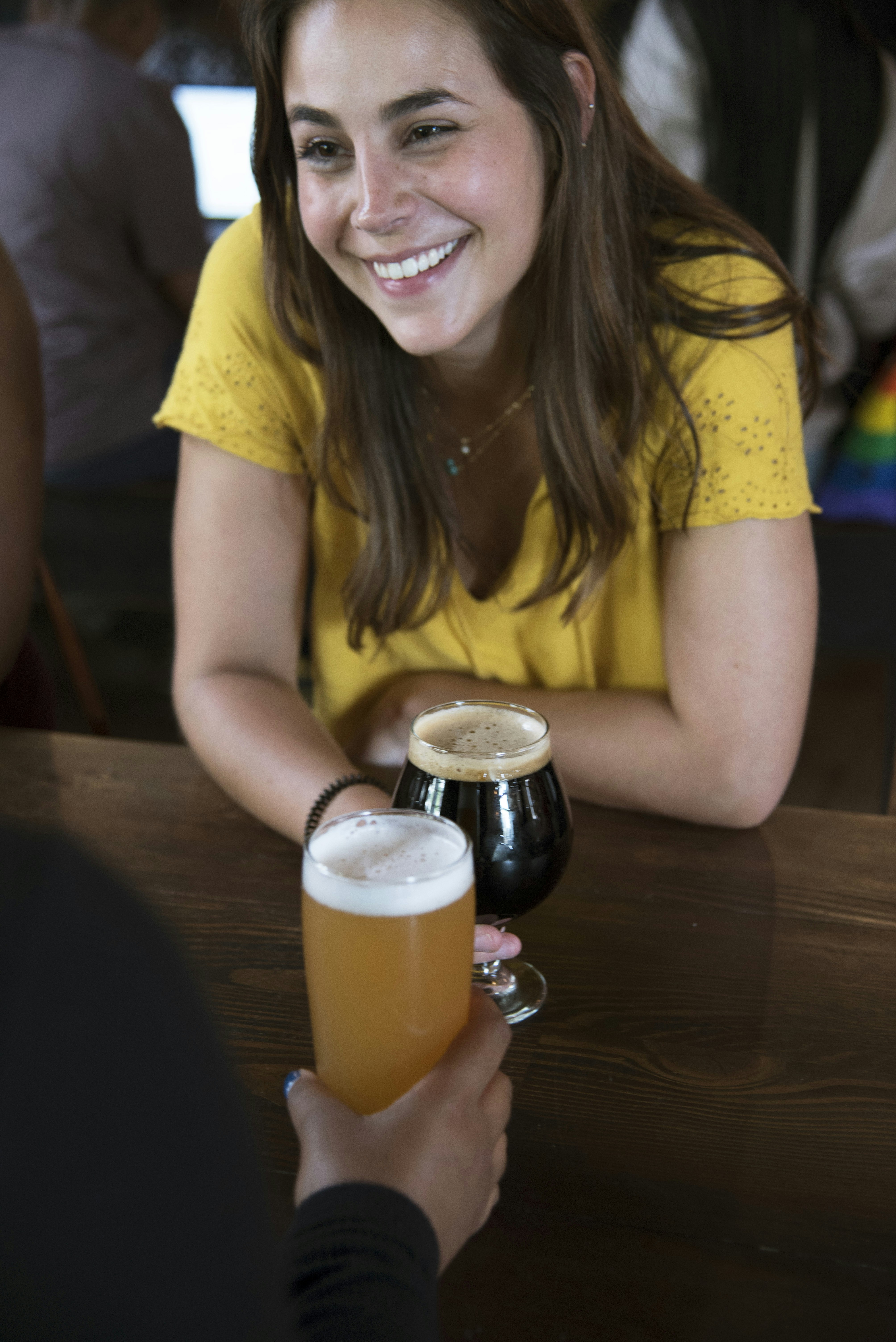Two friends clinking glasses of craft beer in a cozy bar setting, with one smiling brightly in a yellow top.