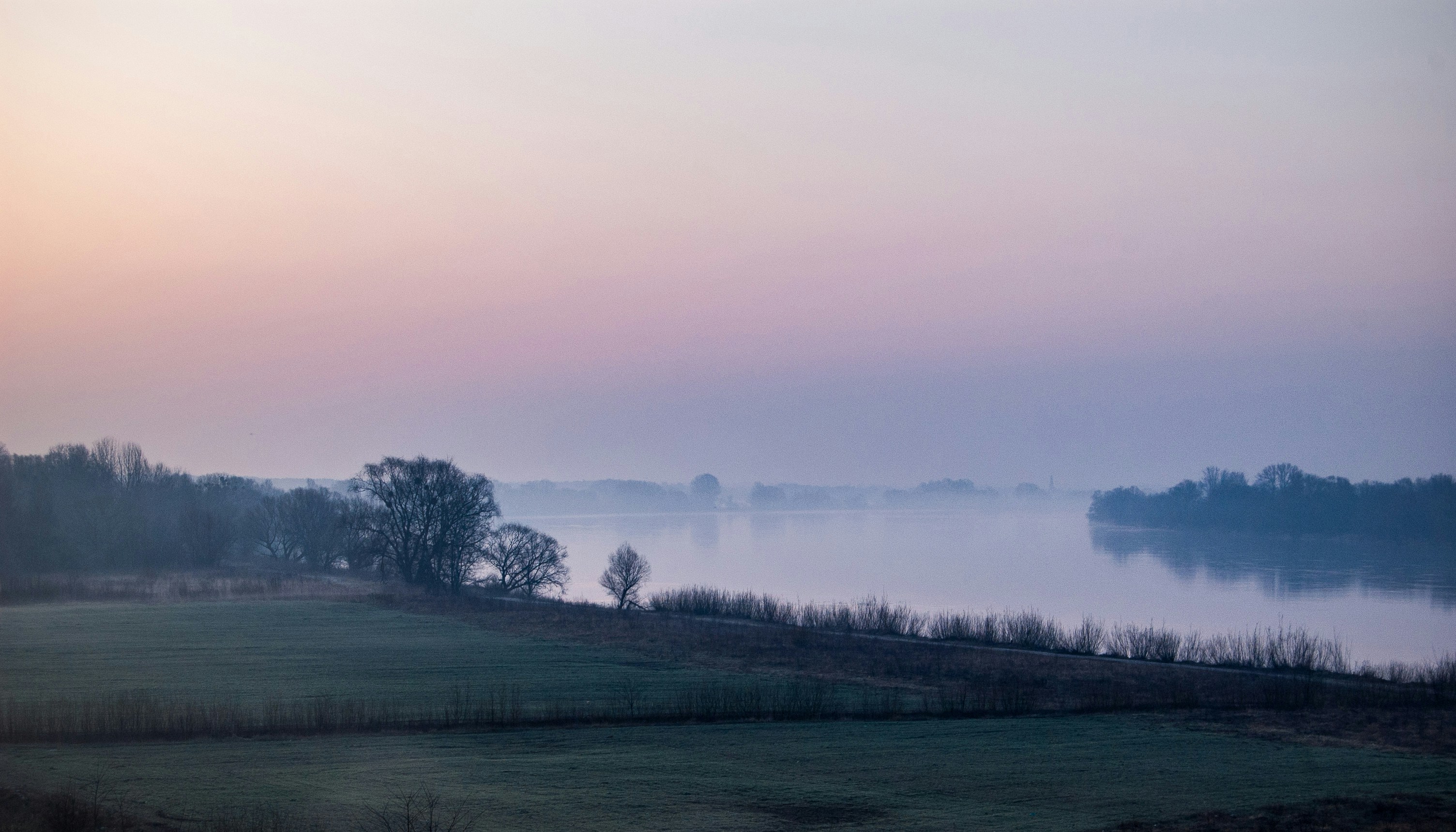 Pastel sunrise over a tranquil river with misty reflections and silhouetted trees.