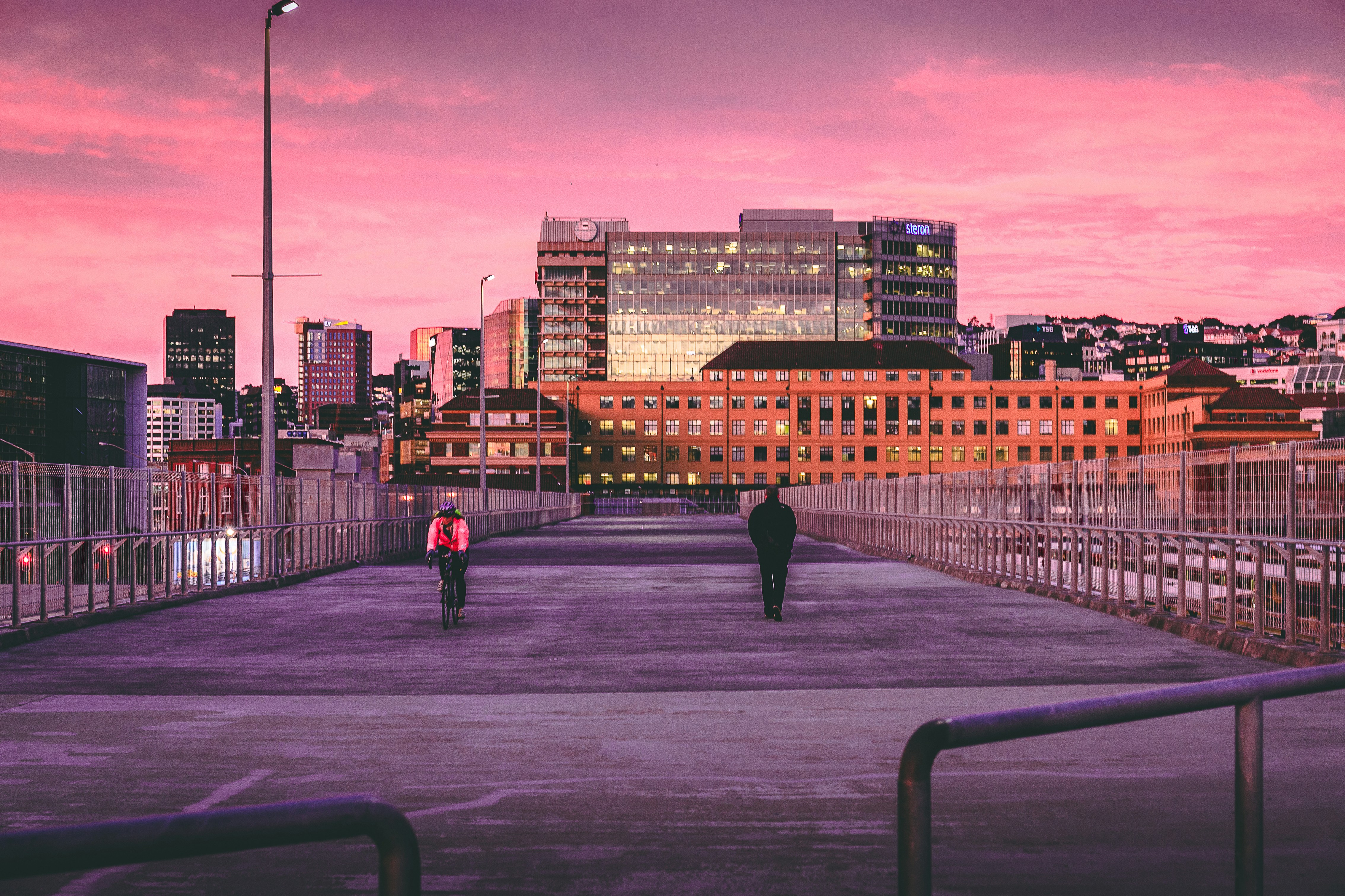 Two people walking on a bridge with a cityscape backdrop under a pink sunset sky.