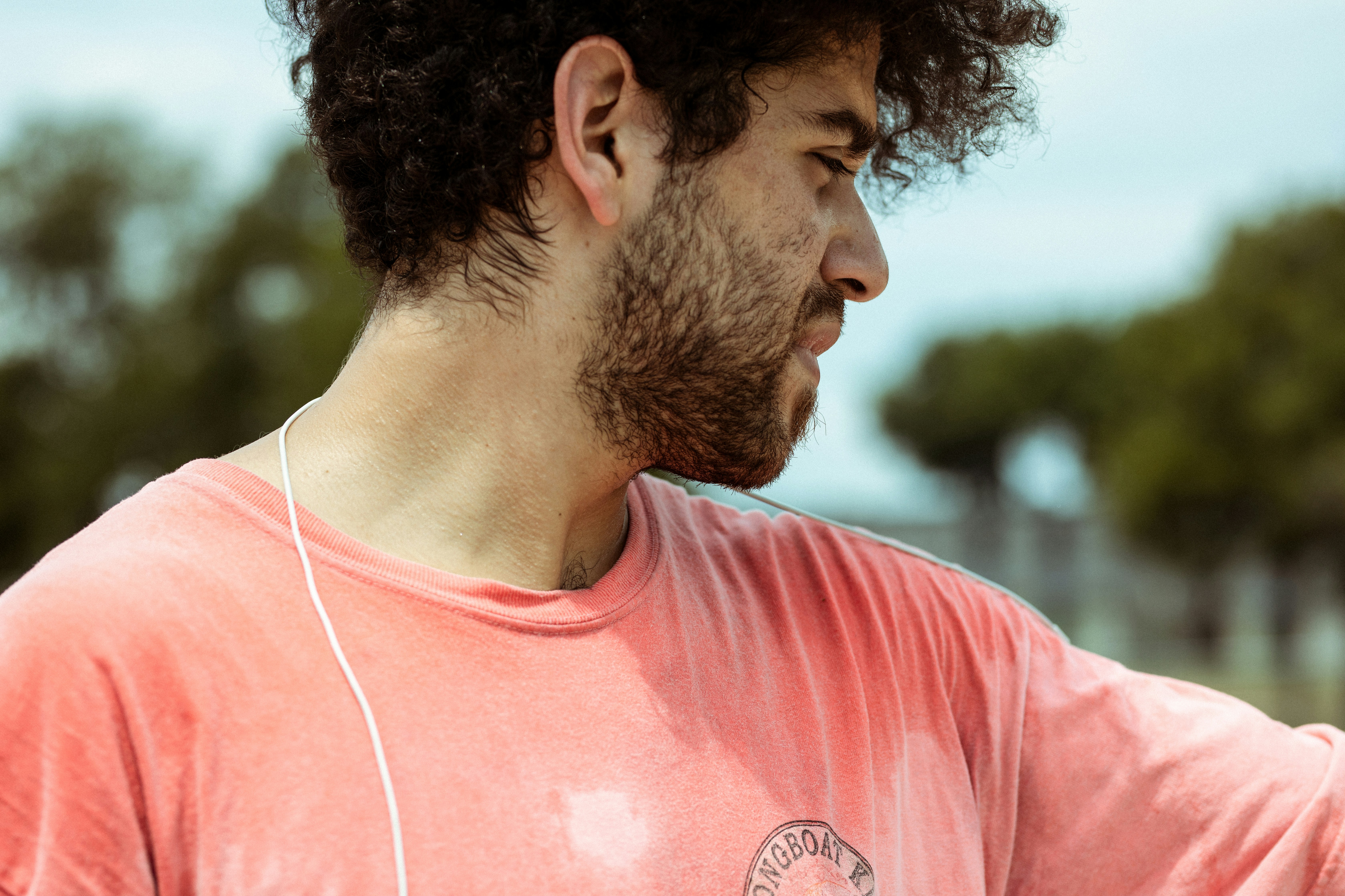 Man in a coral shirt with earbuds looks to the side outdoors.