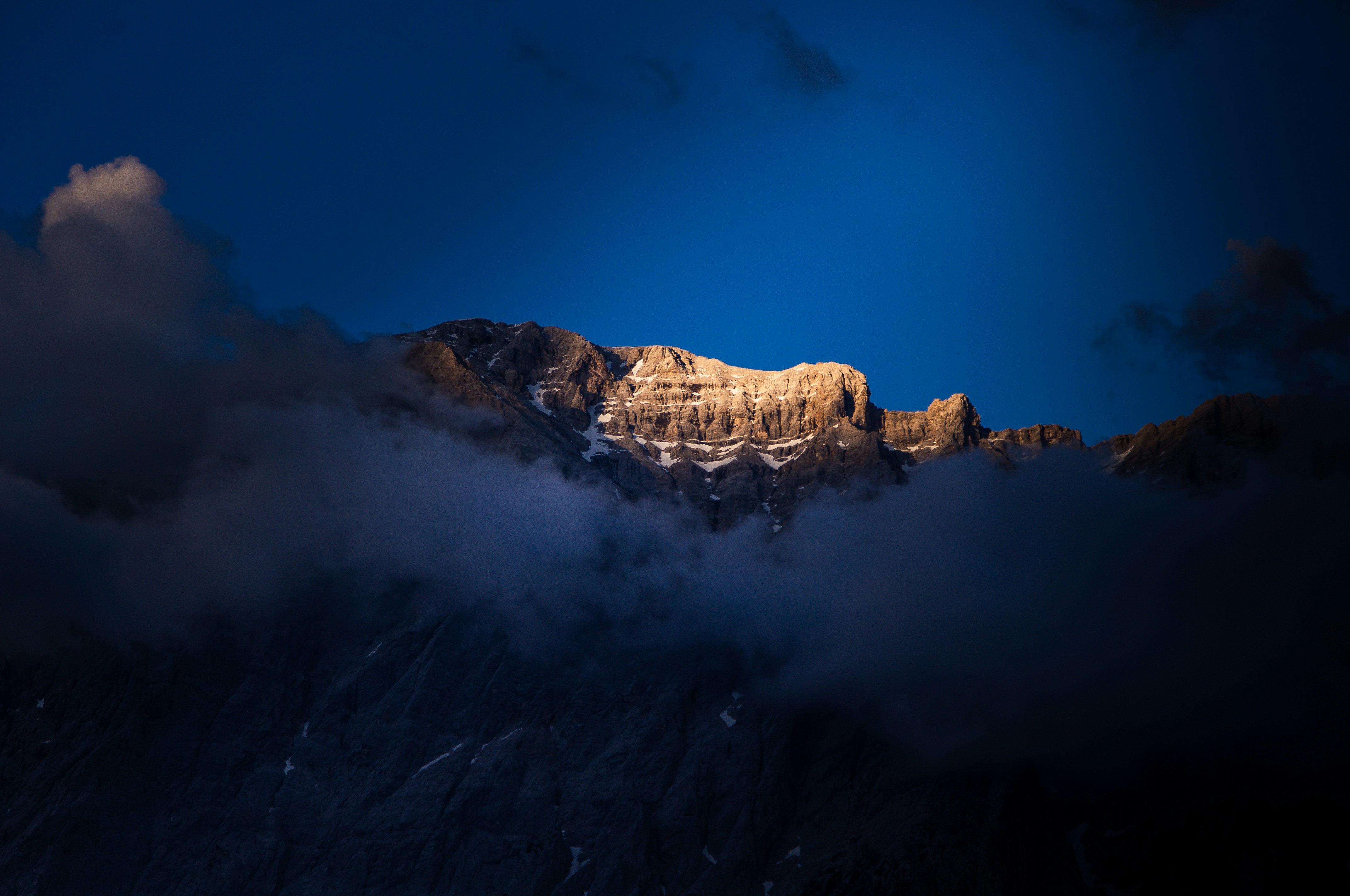 Mountain peak illuminated by warm sunset light, surrounded by dark clouds and deep blue sky.