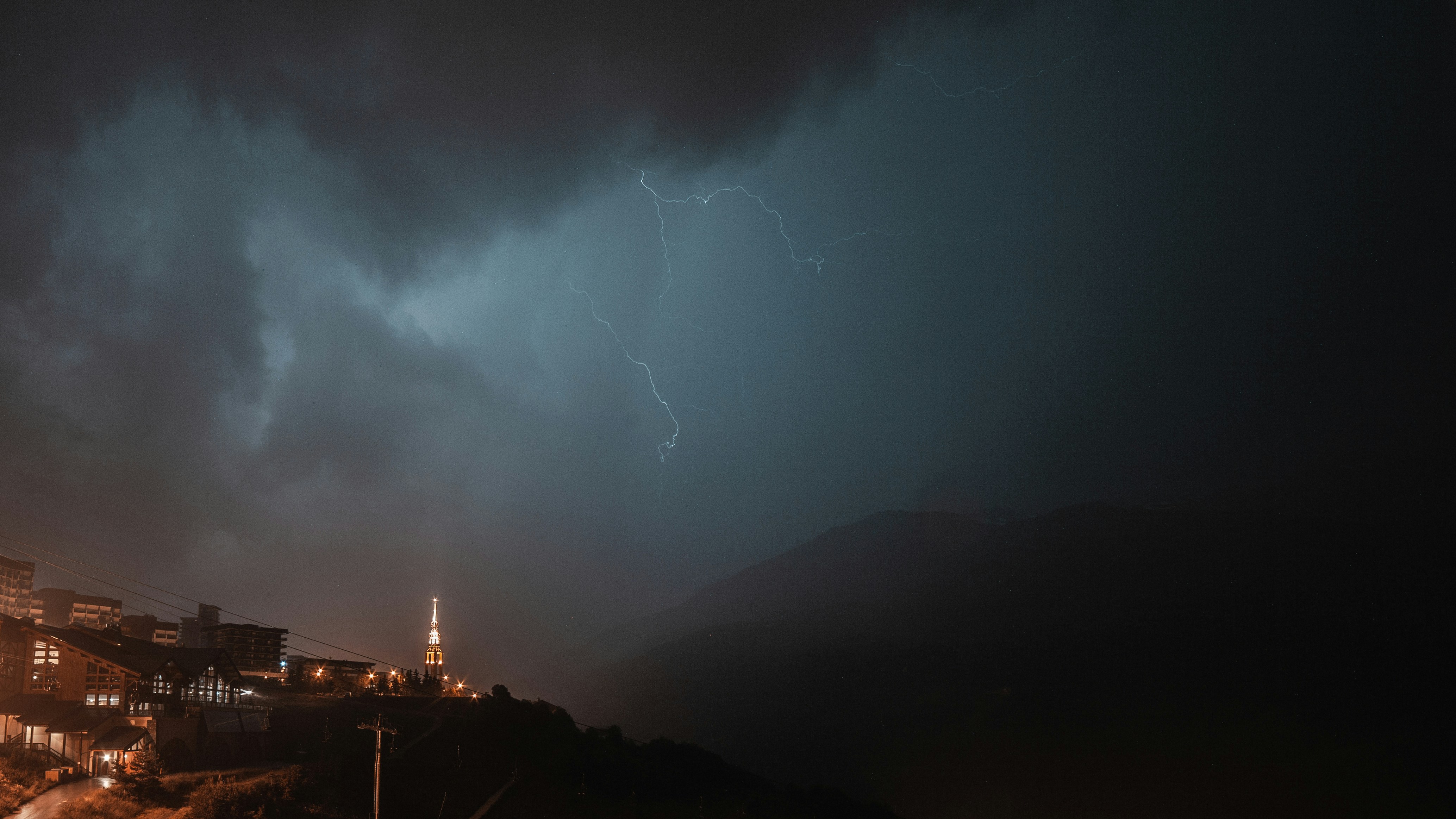 Lightning streaks across a darkened sky above a cityscape, illuminating the buildings below. A dramatic interplay of light and shadow defines the scene.