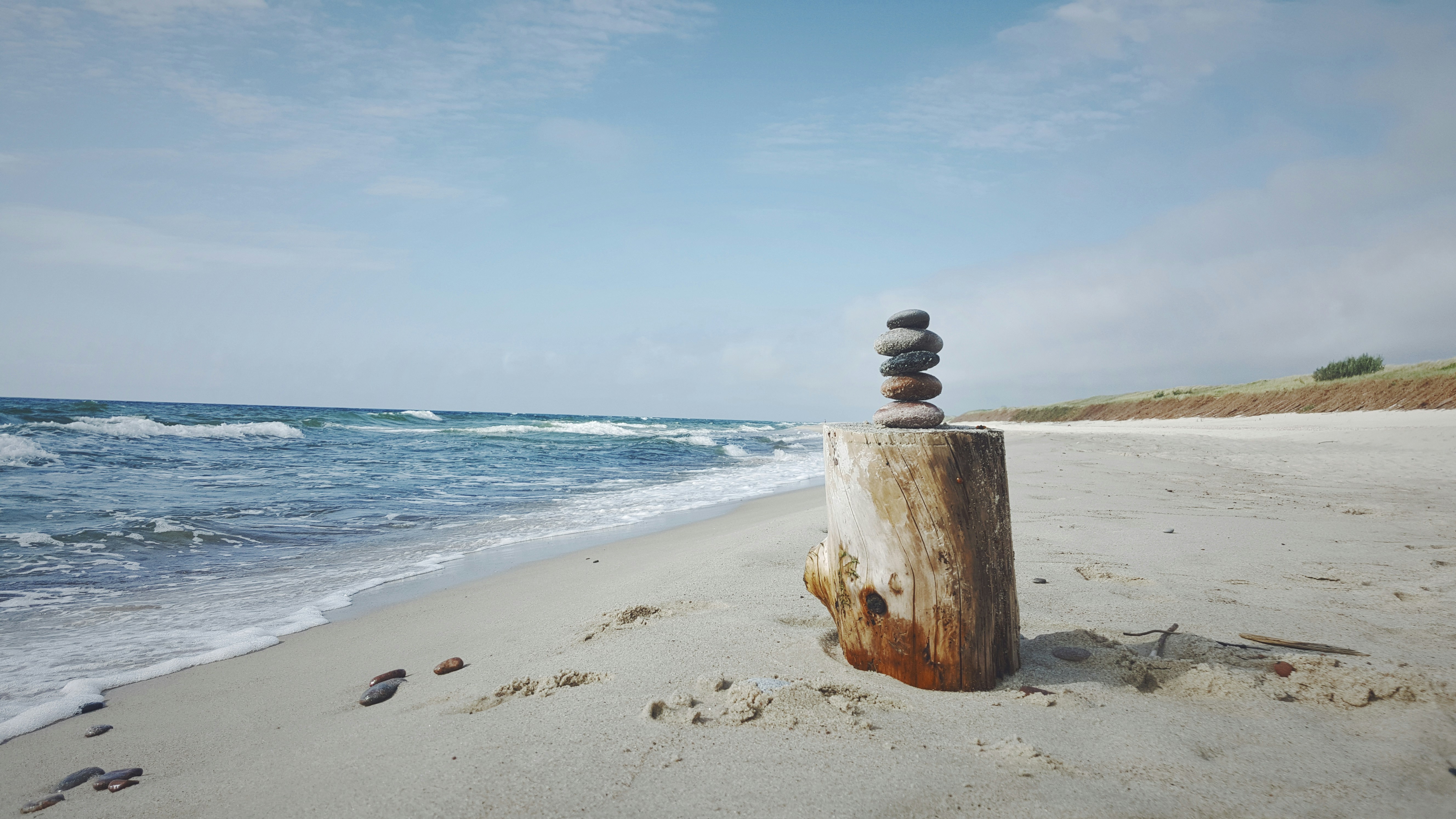 Stack of smooth stones atop a weathered log on a sandy beach with gentle waves in the background.