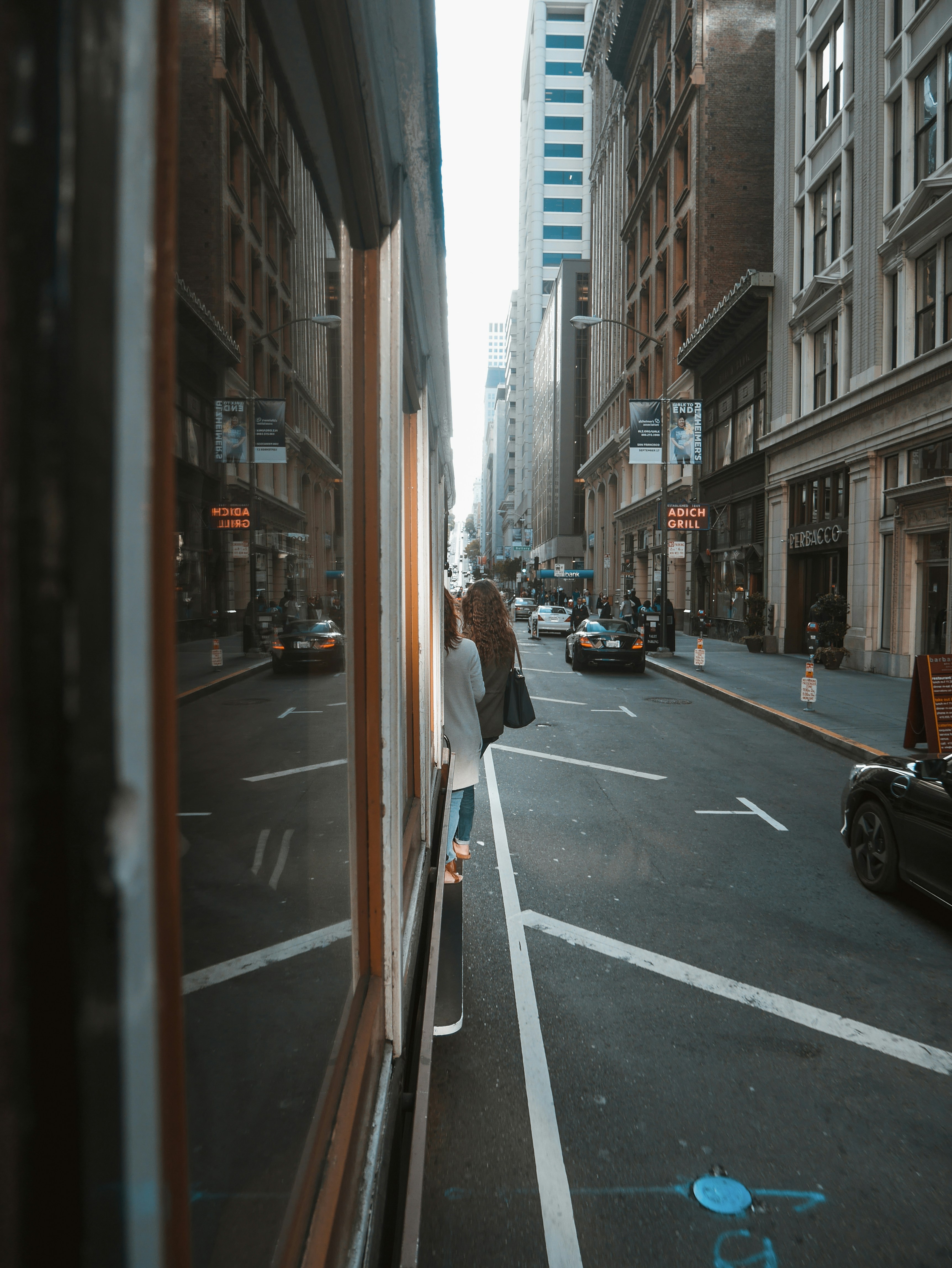 A woman stands beside a window, reflecting the vibrant urban landscape of a bustling street filled with cars and historic architecture.