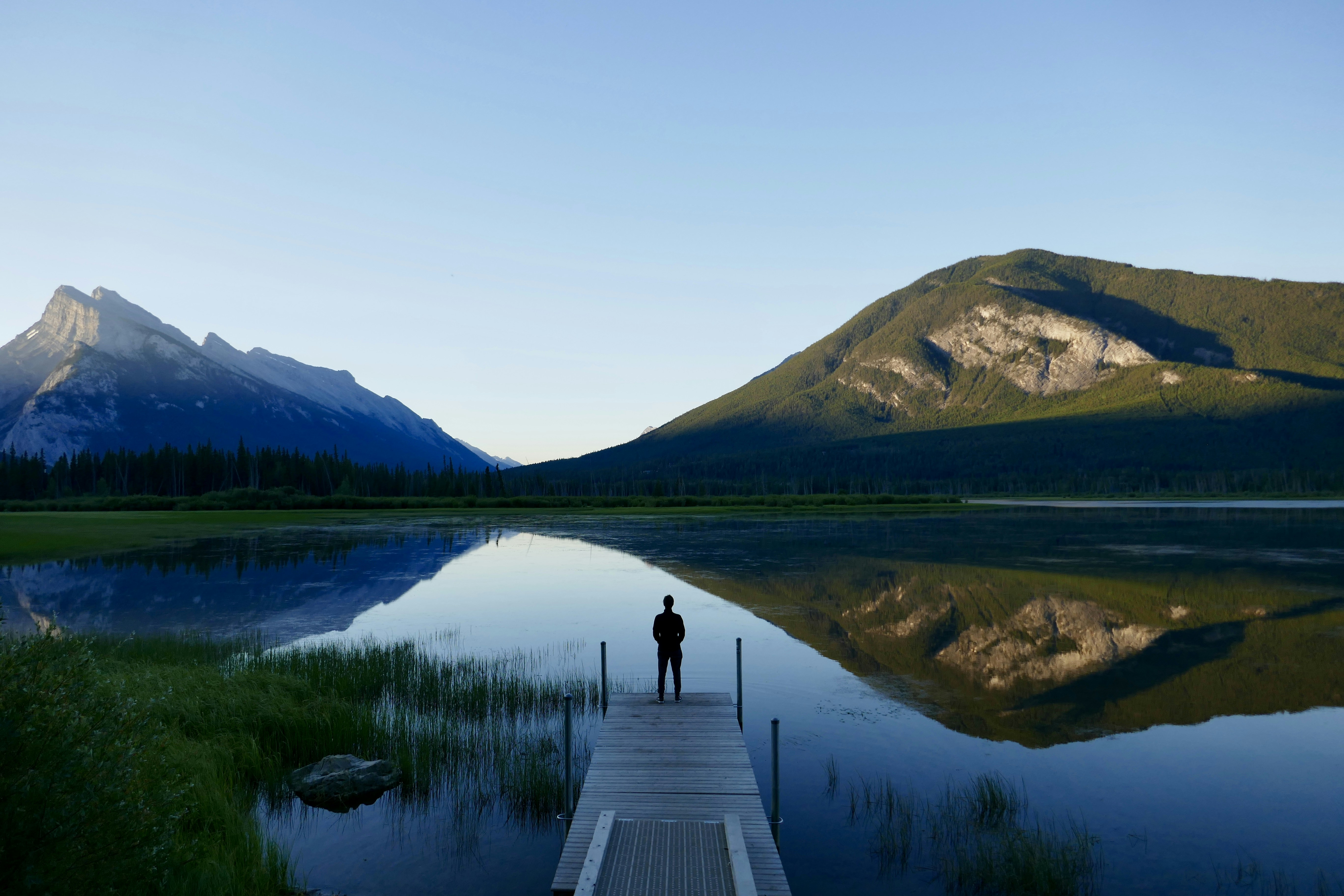 Person standing on a dock reflected in calm lake with mountains in the background under a clear blue sky.