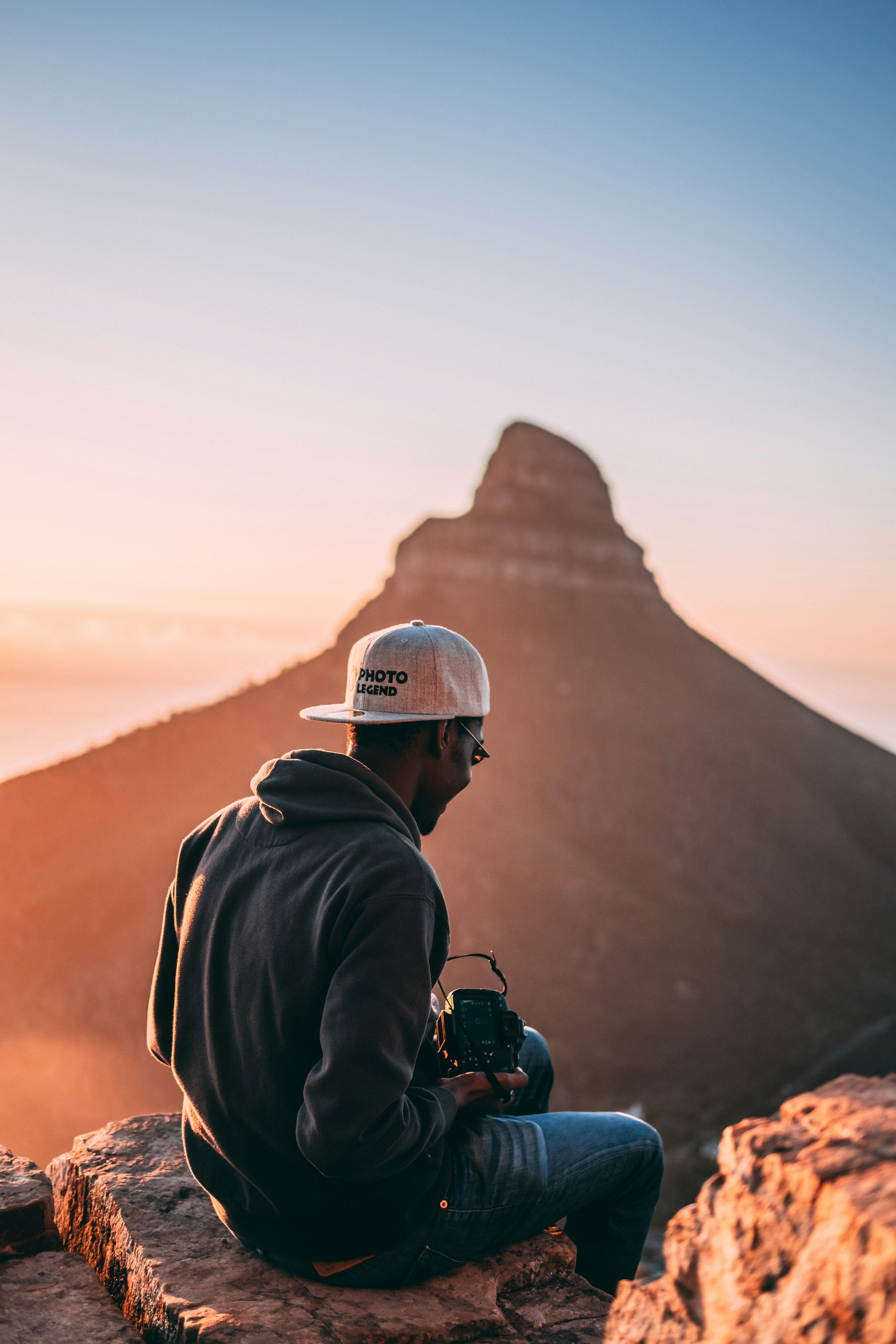 A photographer sits on a rocky ledge, focusing on capturing the serene landscape of a mountain during sunset. The warm light enhances the tranquil atmosphere.