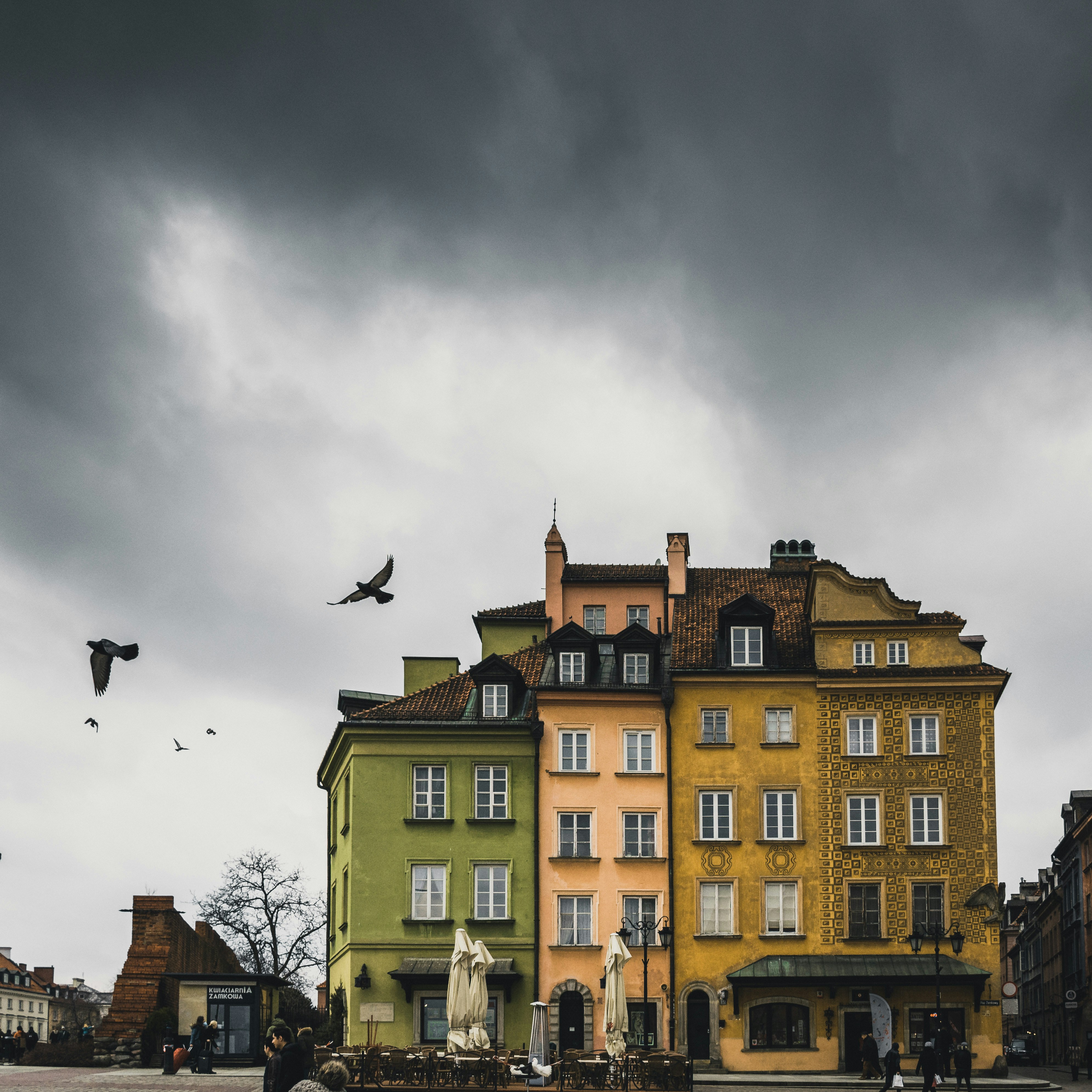 Colorful historic buildings line a square under a dramatic sky, with birds soaring above. A fountain stands as a centerpiece amidst the architecture.