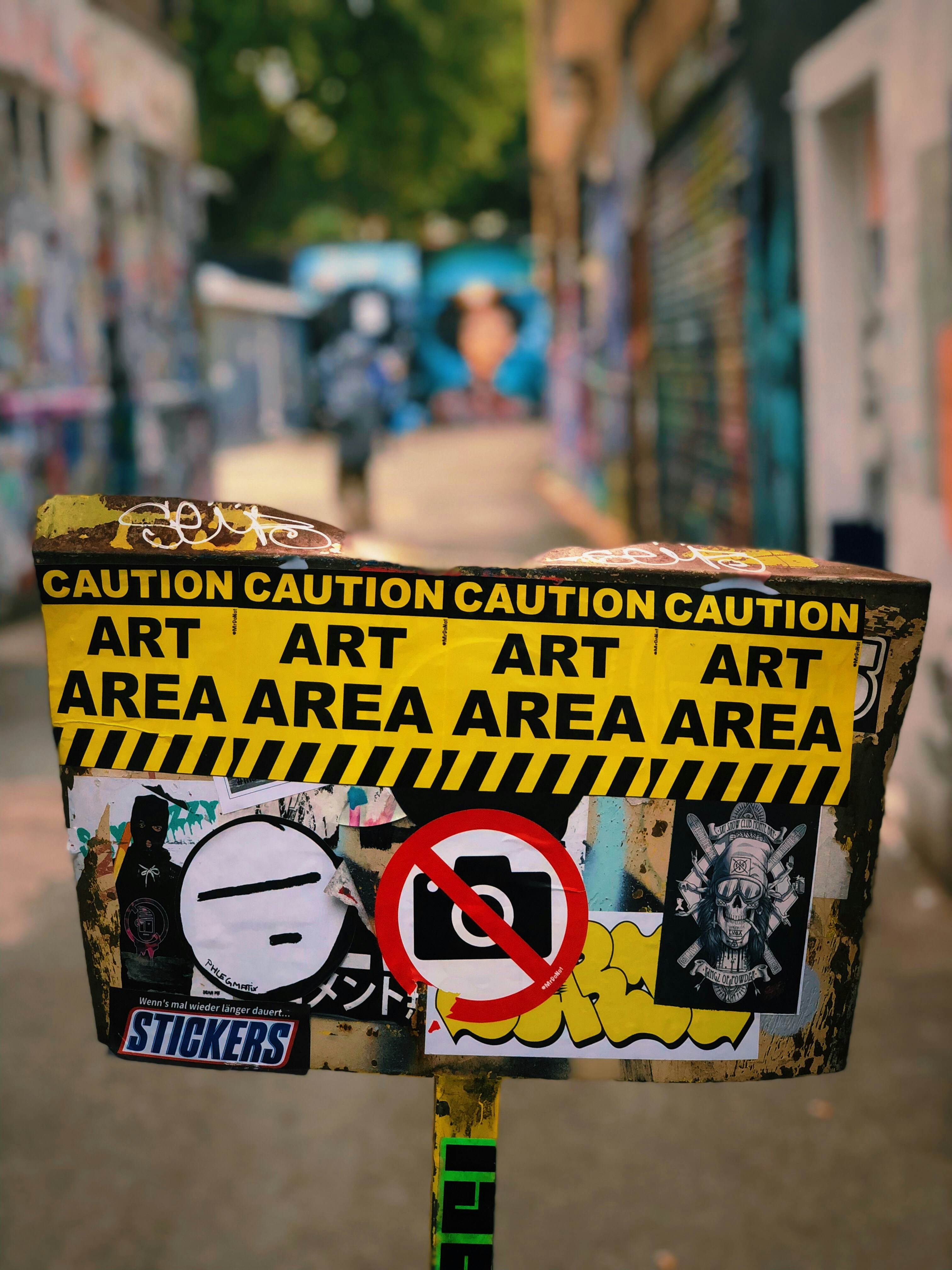 A graffiti-covered sign warning of an art area, adorned with various stickers and illustrations. The vibrant background features a blurred mural, enhancing the urban aesthetic.