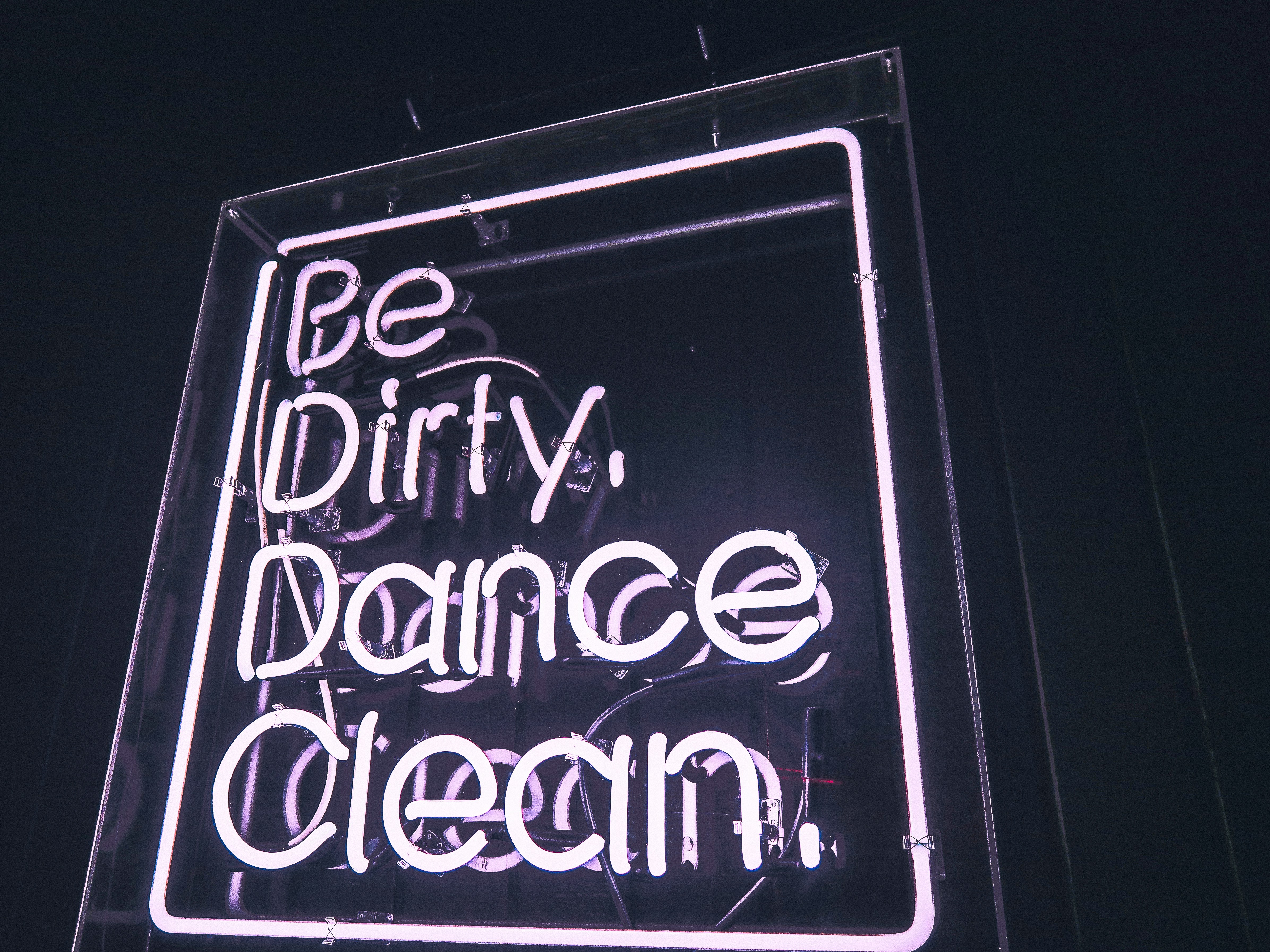 Neon sign with the words 'Be Dirty, Dance Clean' glowing against a dark background.