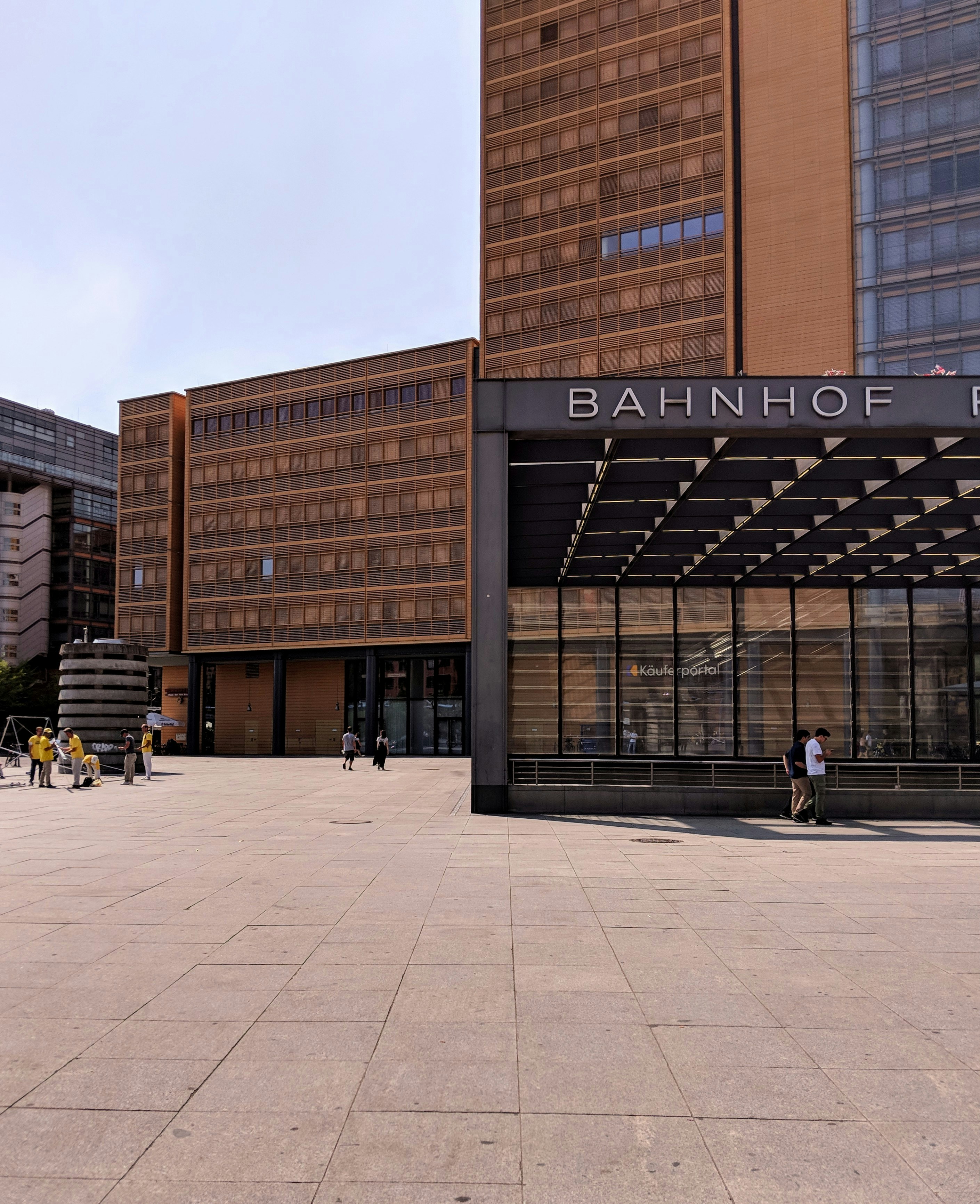 Contemporary train station facade with large glass windows and bold architectural lines, bustling with people in a vibrant urban setting.