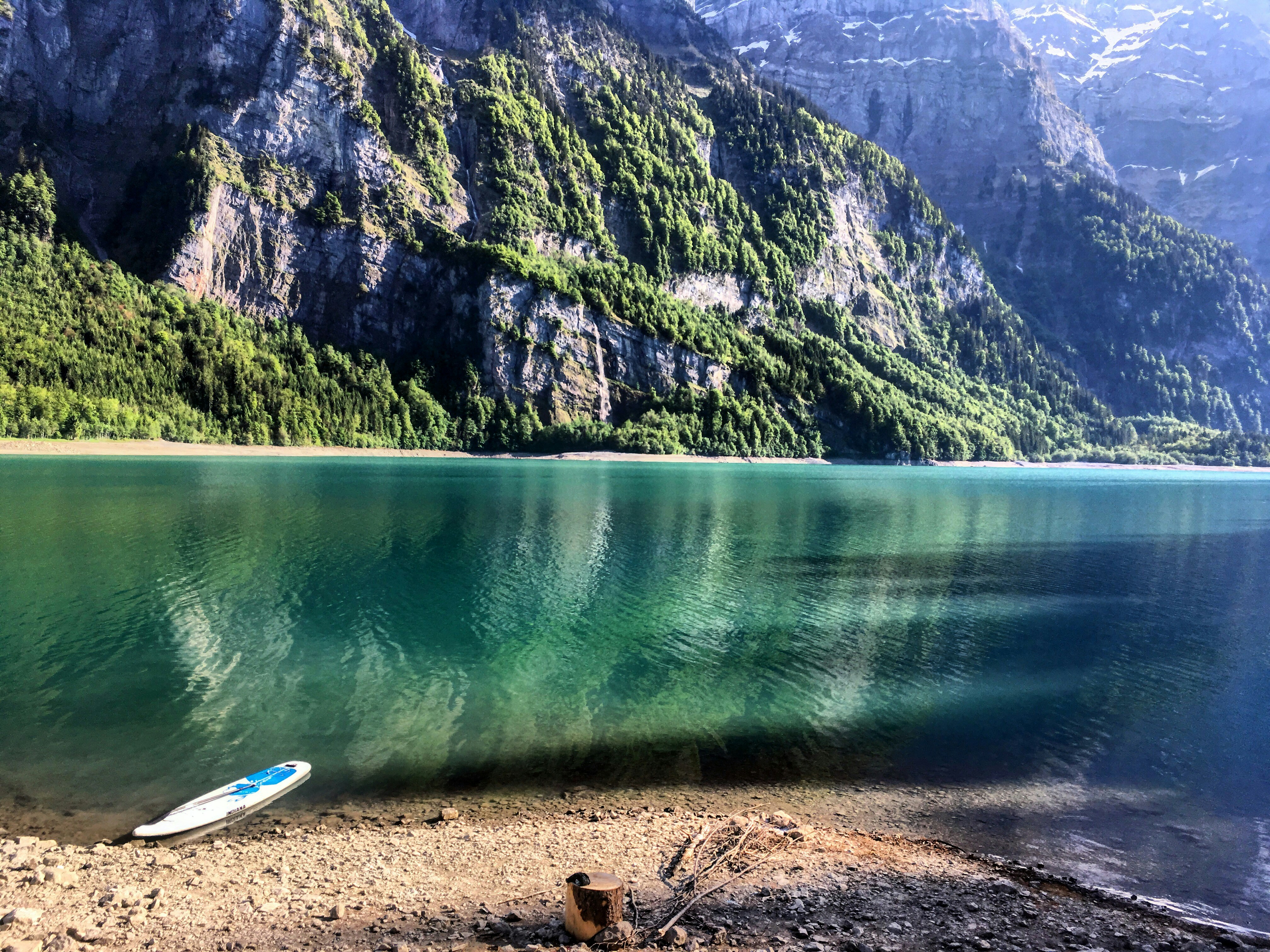 Paddleboard resting on the shore of a serene lake surrounded by towering, forested cliffs under a clear sky.