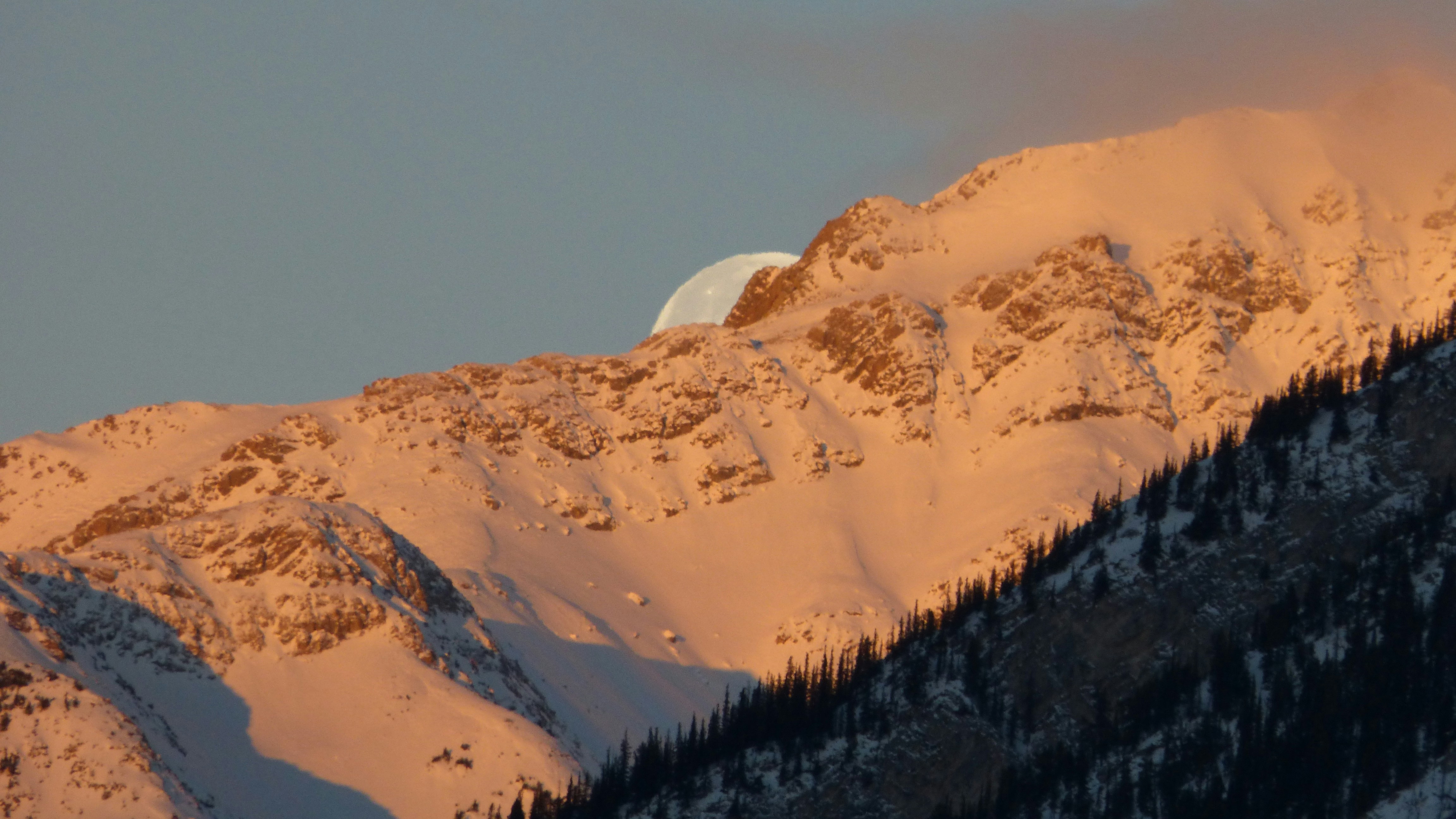 Moon setting behind sunlit snow-capped mountains in Banff National Park.