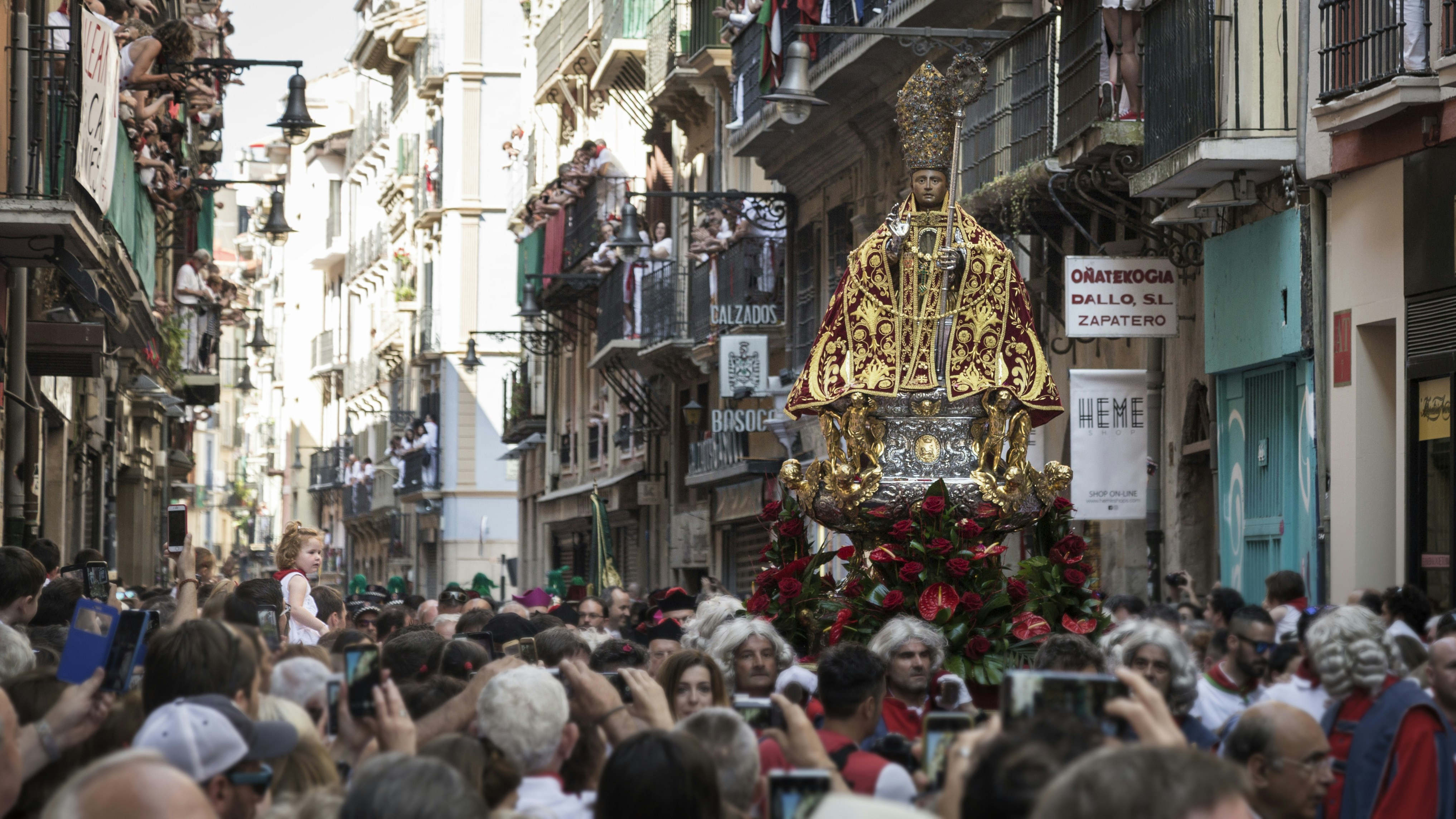 Religious statue adorned with gold and flowers parading through a crowded, narrow street.