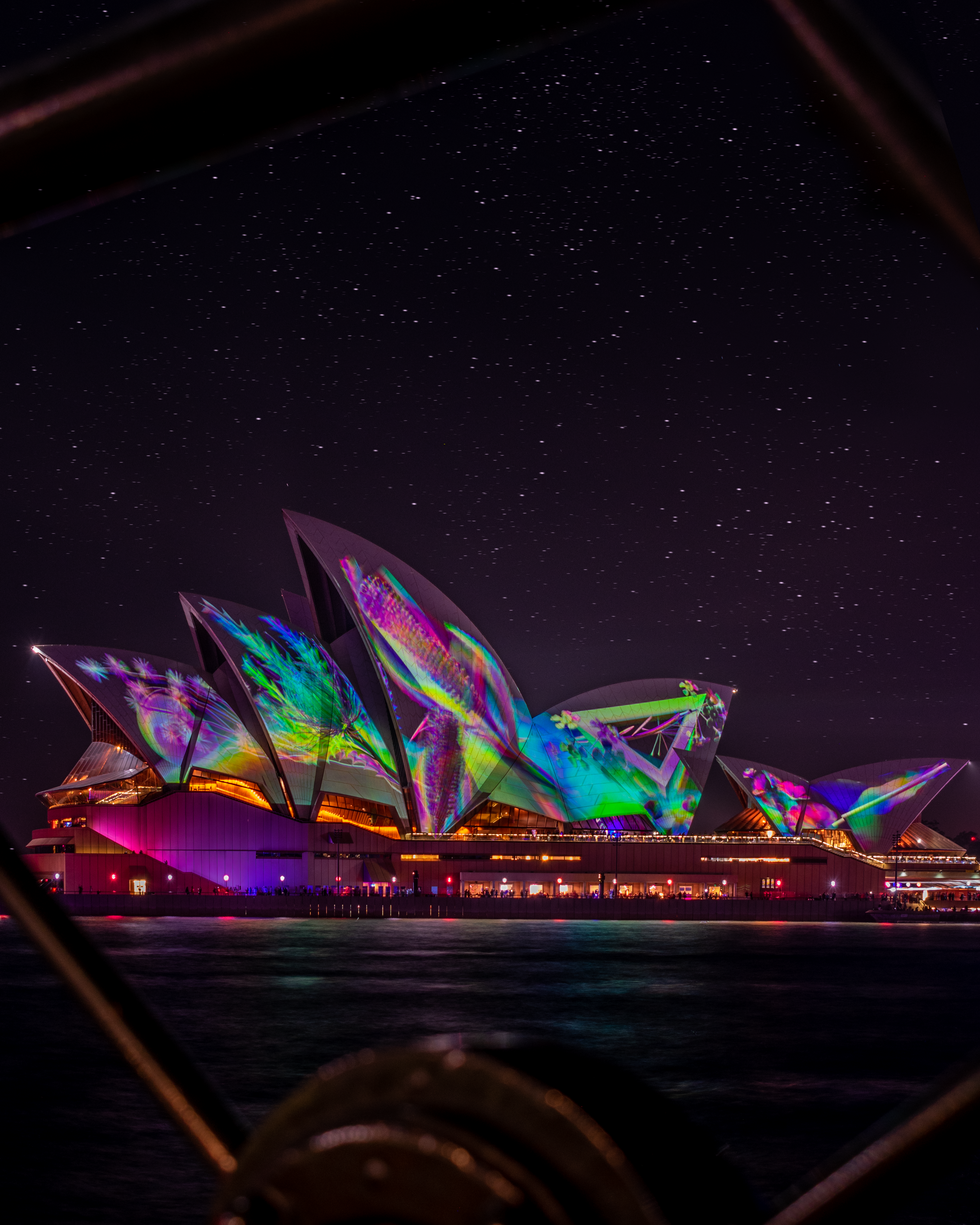 Night photograph of the Sydney Opera House illuminated by colorful projection on its sails, with a star-filled sky and reflections on the water.
