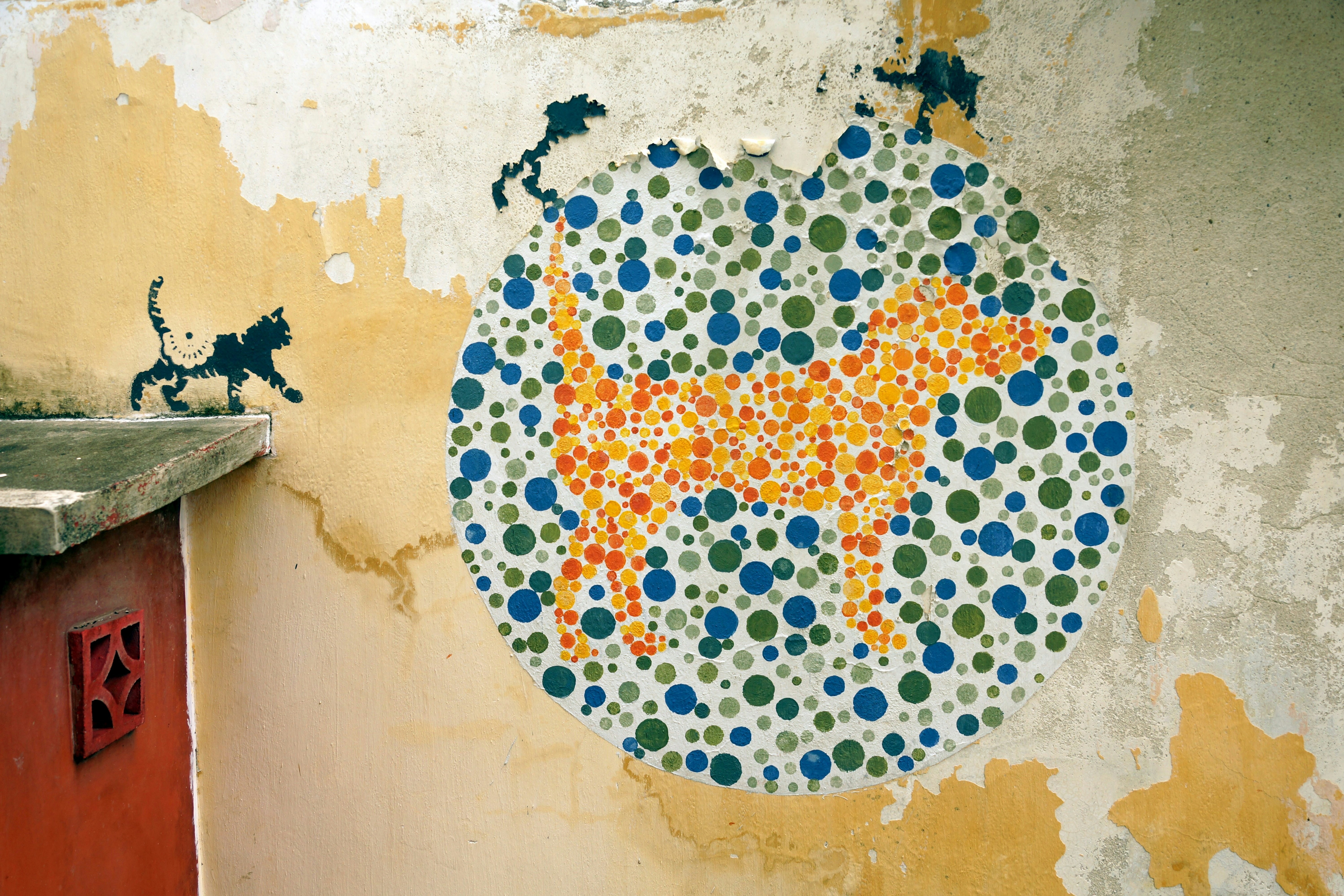 Colorful mosaic of a dog surrounded by vibrant dots on a weathered wall, complemented by a silhouette of a cat in the upper left corner.