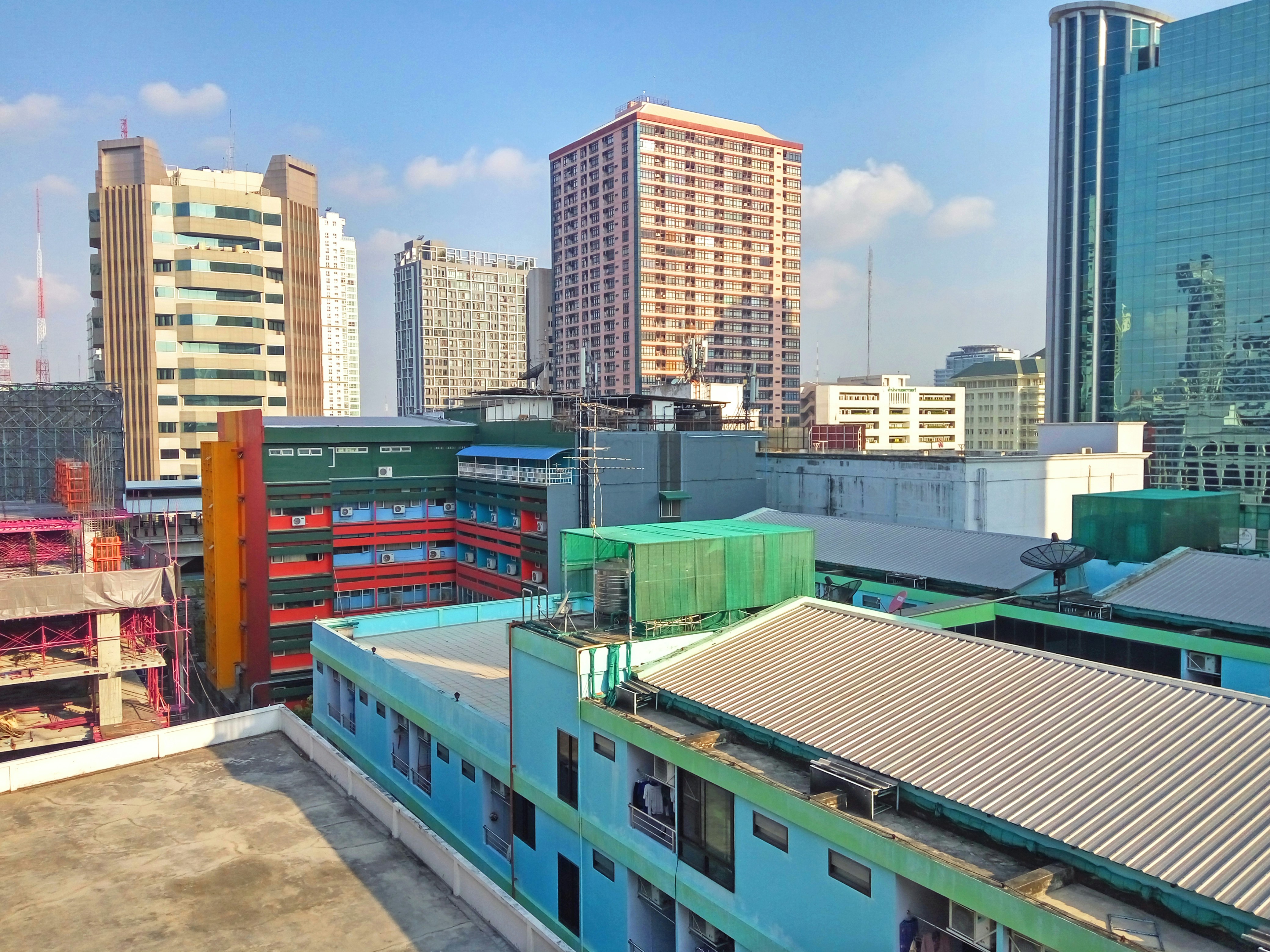 A rooftop vantage captures teal roofs and a dense city skyline under bright daylight. The scene highlights architectural contrasts between colorful low-rise structures and tall glass towers.