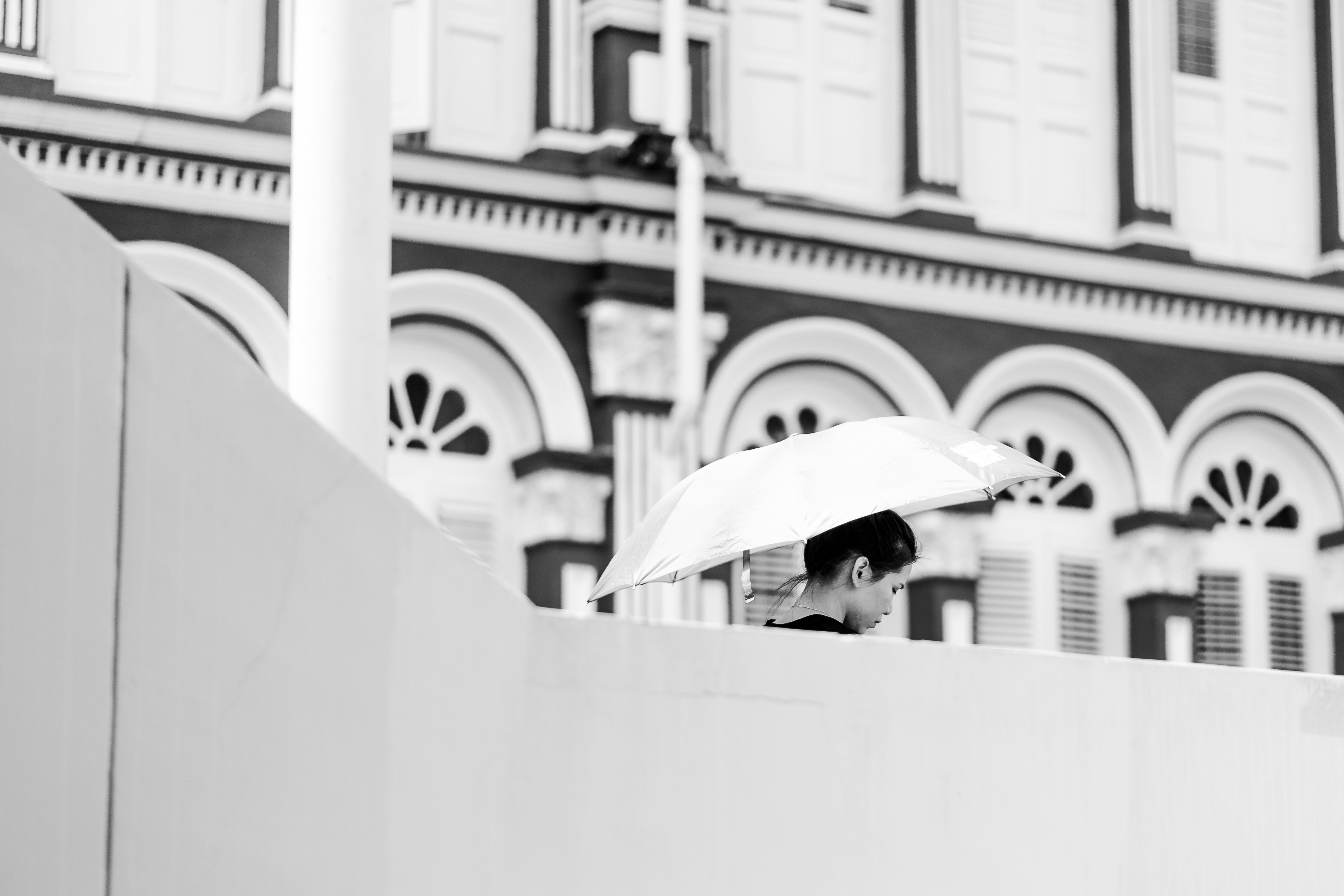 A person holding a white umbrella walks beside a stark white wall, creating a contrast against the dark architectural backdrop.