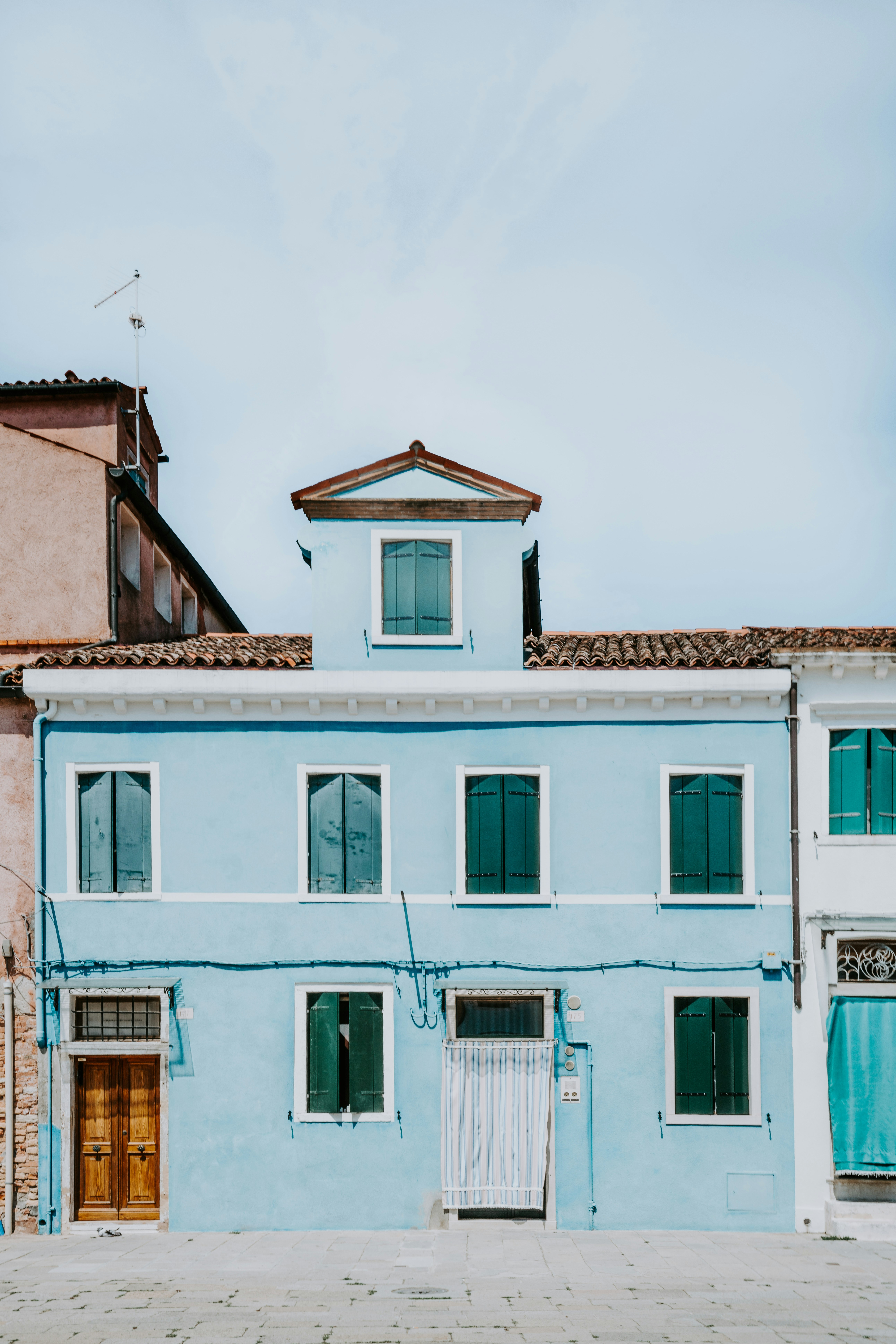 This image captures a charming blue facade of a traditional building, beautifully framed against a clear, expansive sky. The composition highlights the symmetrical arrangement of windows, each adorned with dark shutters that contrast with the pastel blue walls. The soft lighting and muted color palette create a tranquil atmosphere, making the scene visually striking and inviting.