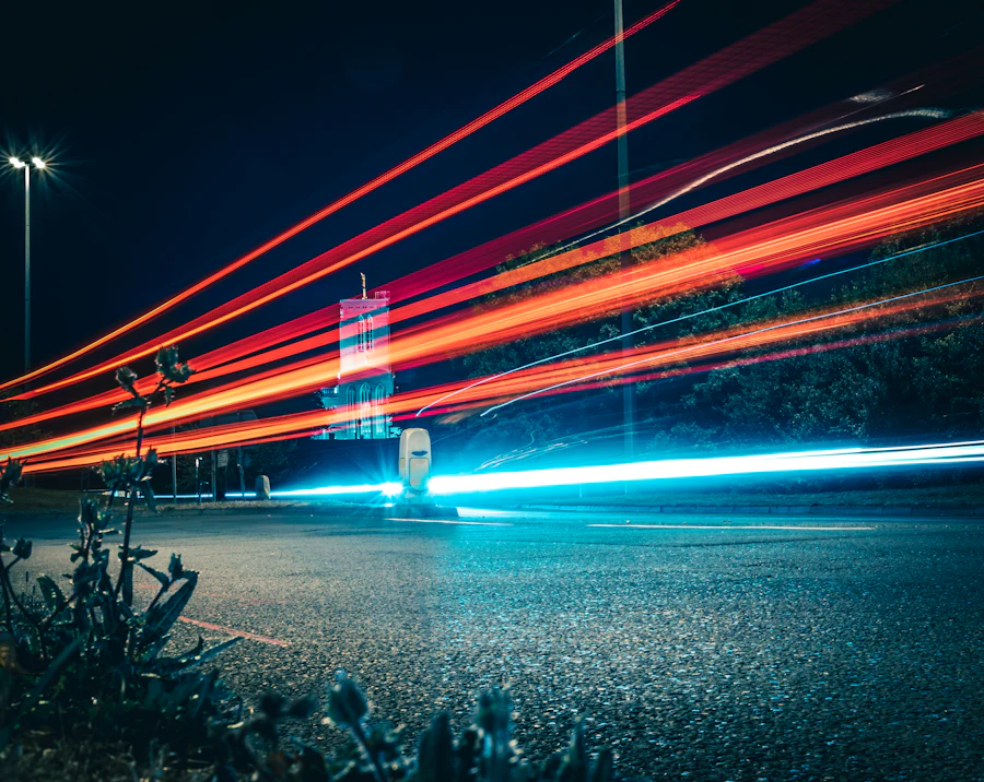 Long-exposure light trails streak across a dark road at night, with orange-red lines overhead and a bright blue line near the pavement. A small illuminated structure and roadside plants sit in the foreground.