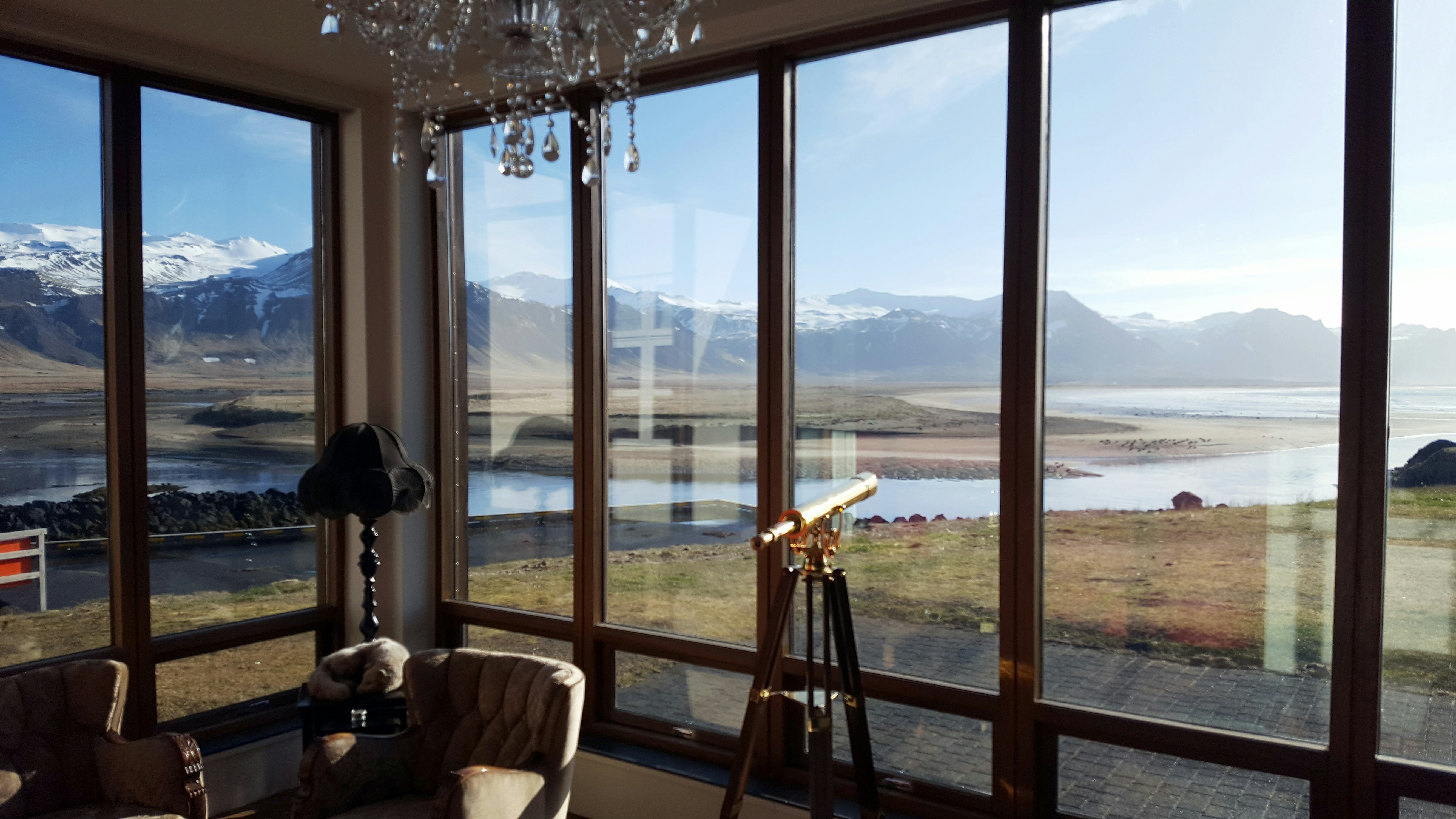 Cozy interior scene featuring large windows framing a breathtaking view of mountains and a tranquil body of water, with a telescope positioned for observation.