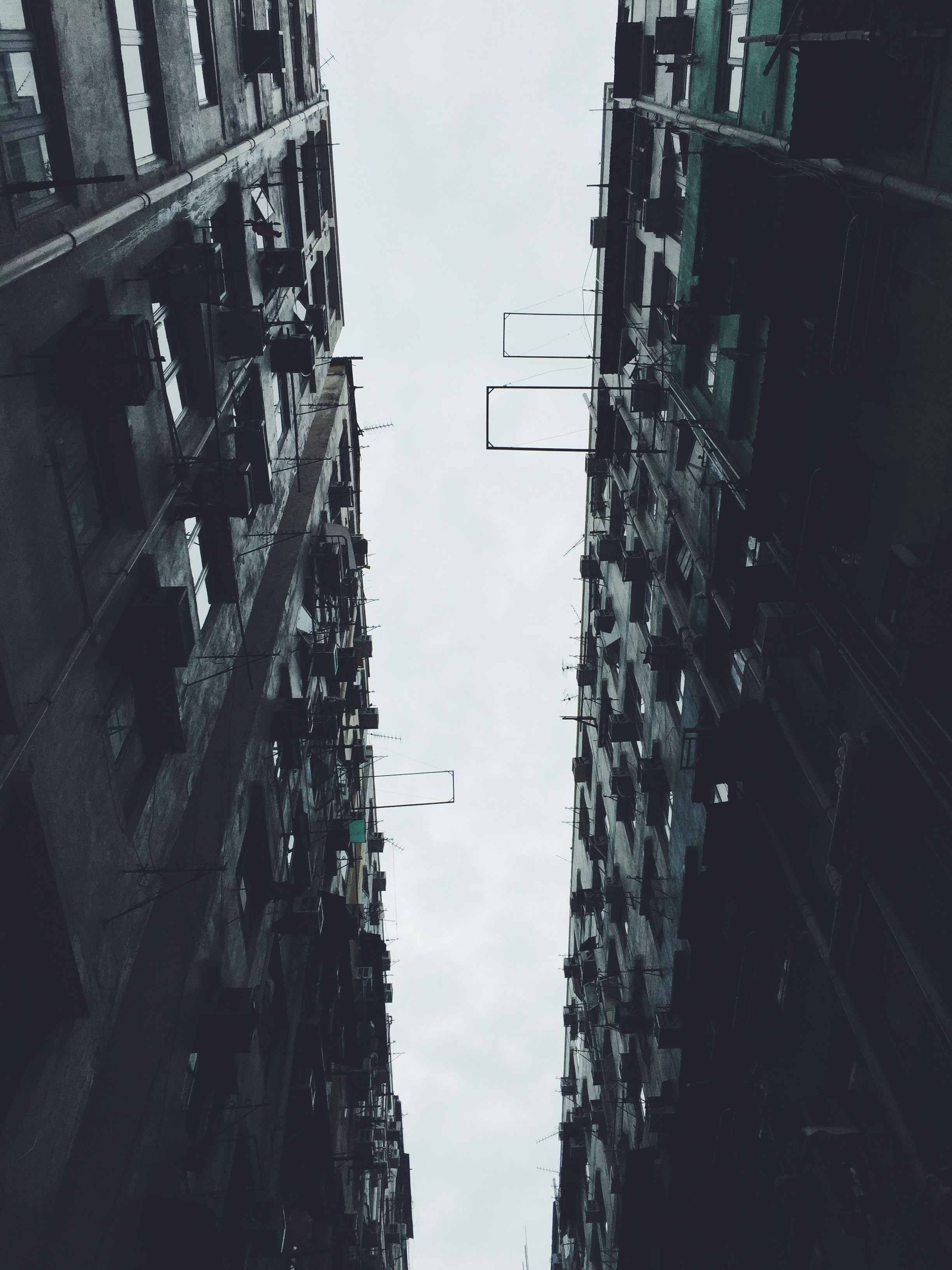 Narrow alleyway between tall, weathered buildings, showcasing a cloudy sky above and urban elements like air conditioning units and antennas.