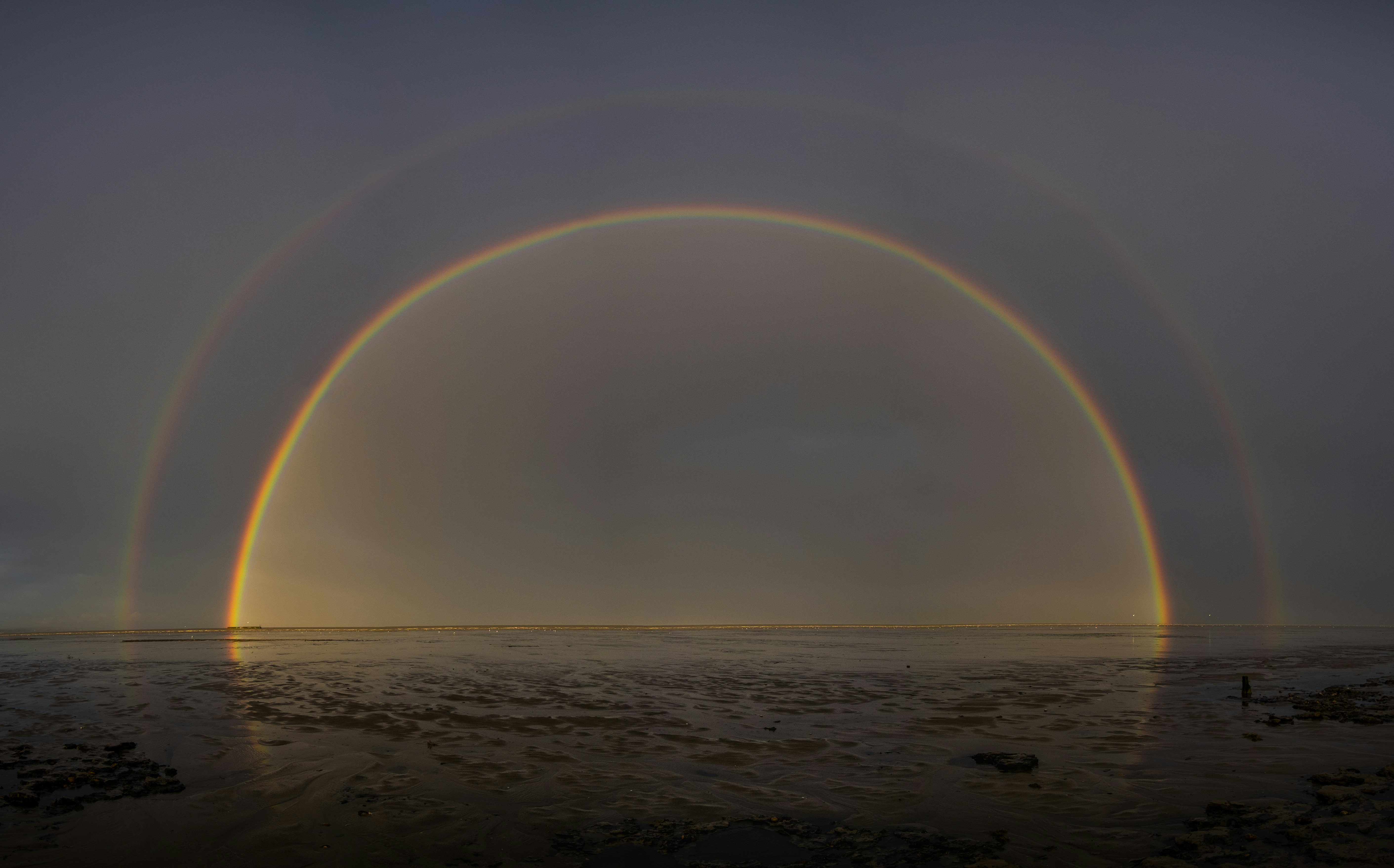 A vibrant double rainbow arches over a tranquil shoreline, framed by a moody sky. The scene captures the interplay of light and nature's beauty.