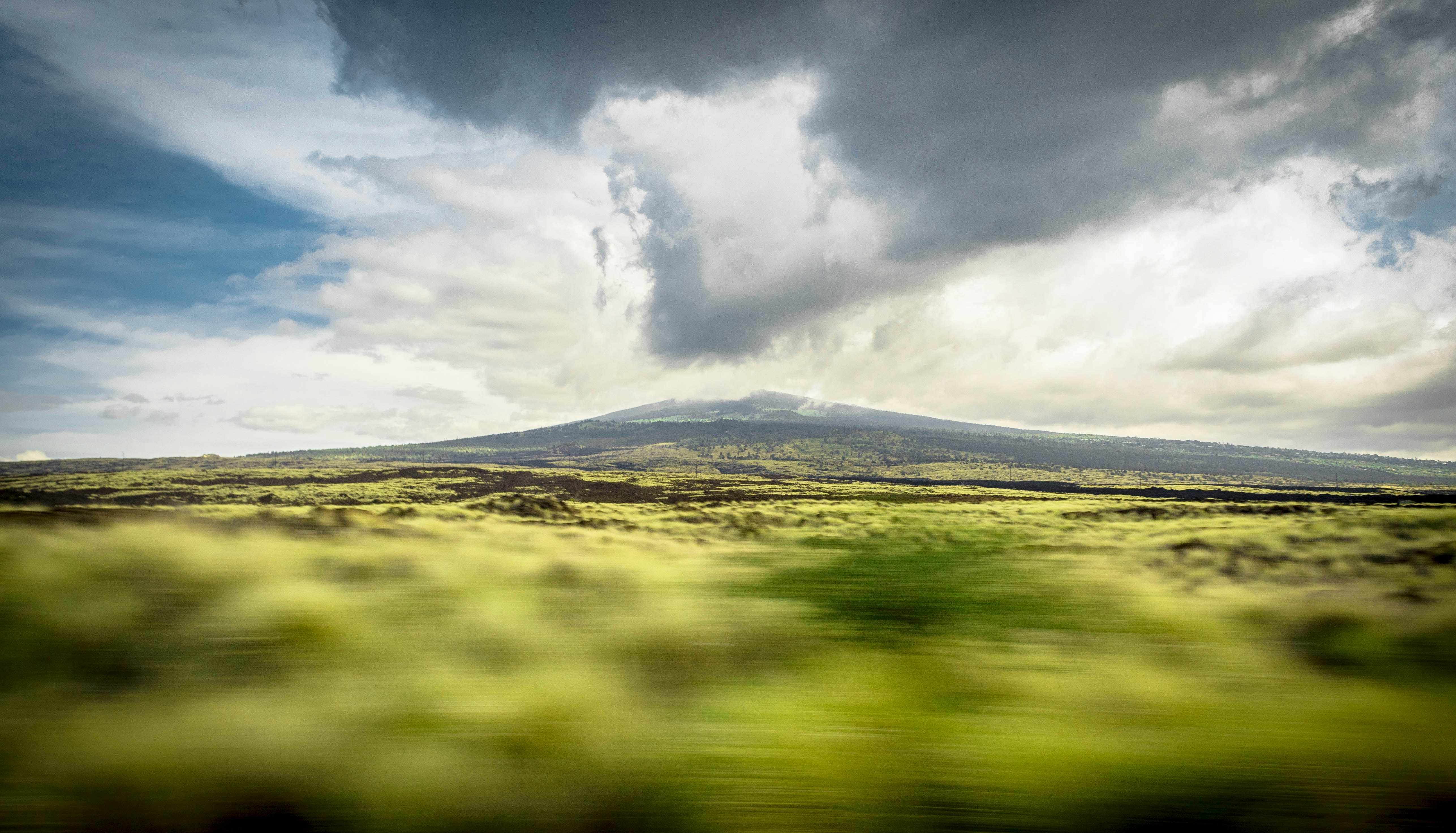Vast green landscape with a distant mountain shrouded in clouds, creating a dynamic interplay of light and shadow.