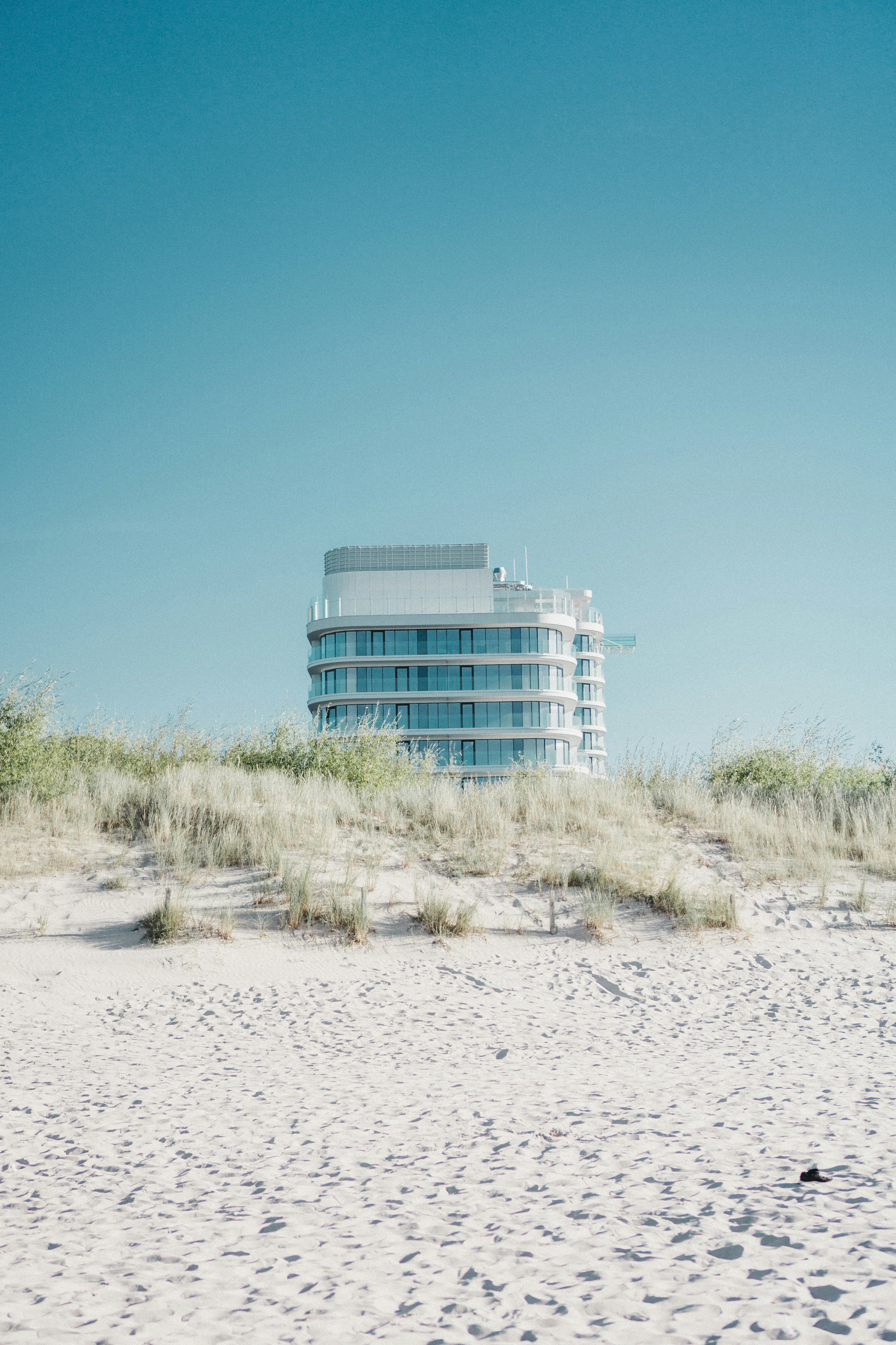 A contemporary building rises above sandy dunes, framed by lush greenery under a clear blue sky.