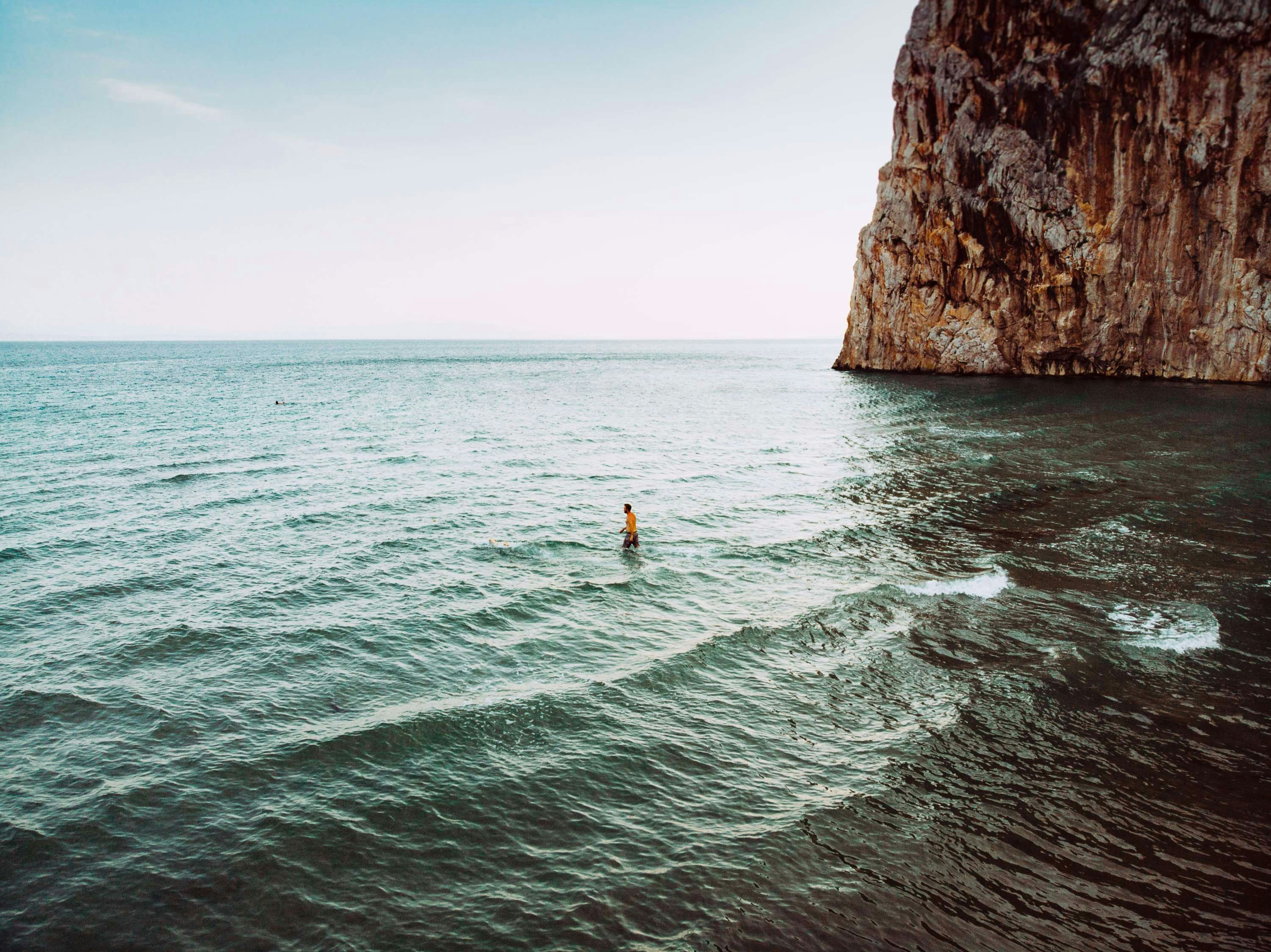 Person wading in calm ocean near a towering rocky cliff.