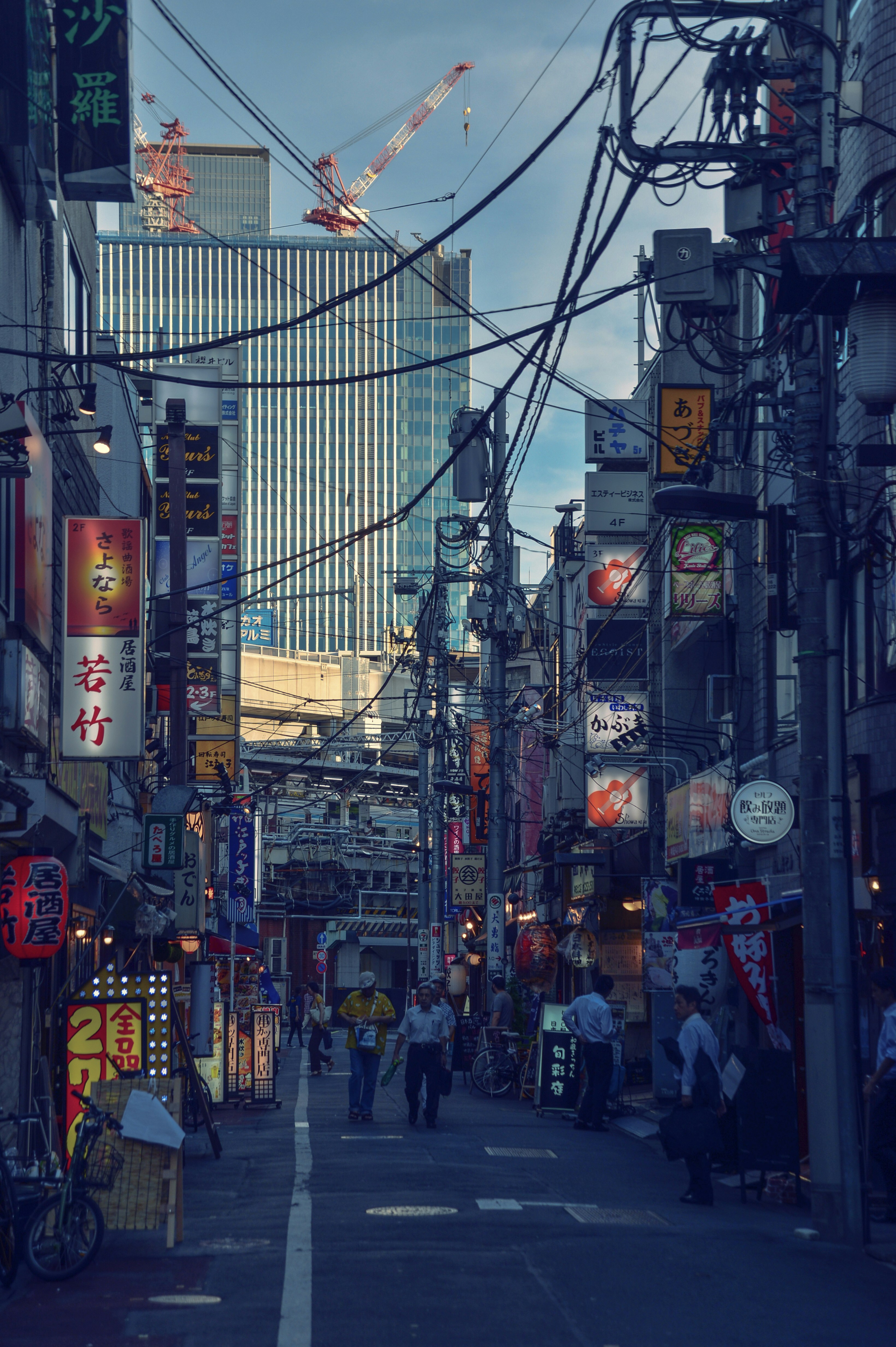 Narrow street lined with vibrant signage and bustling pedestrians, framed by towering skyscrapers in the background. A blend of old and new urban life.